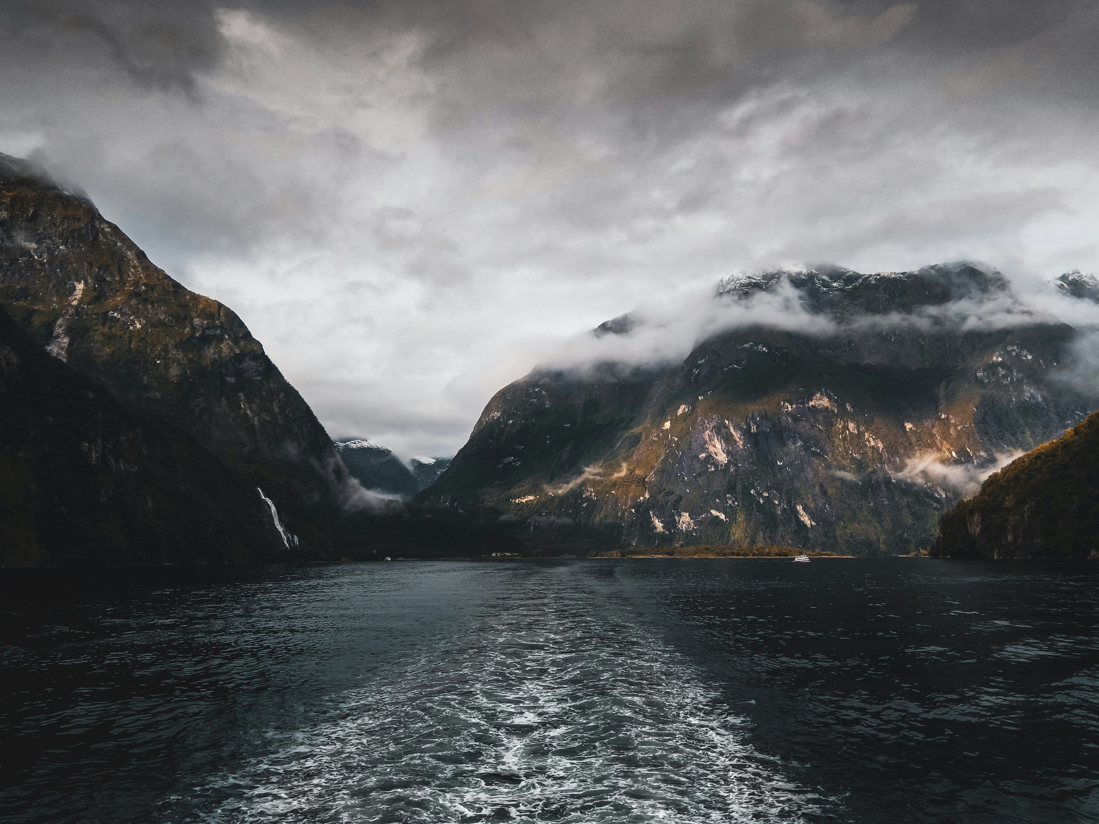 Cloud-kissed mountains rise over dark waters with a wake trailing behind a boat.