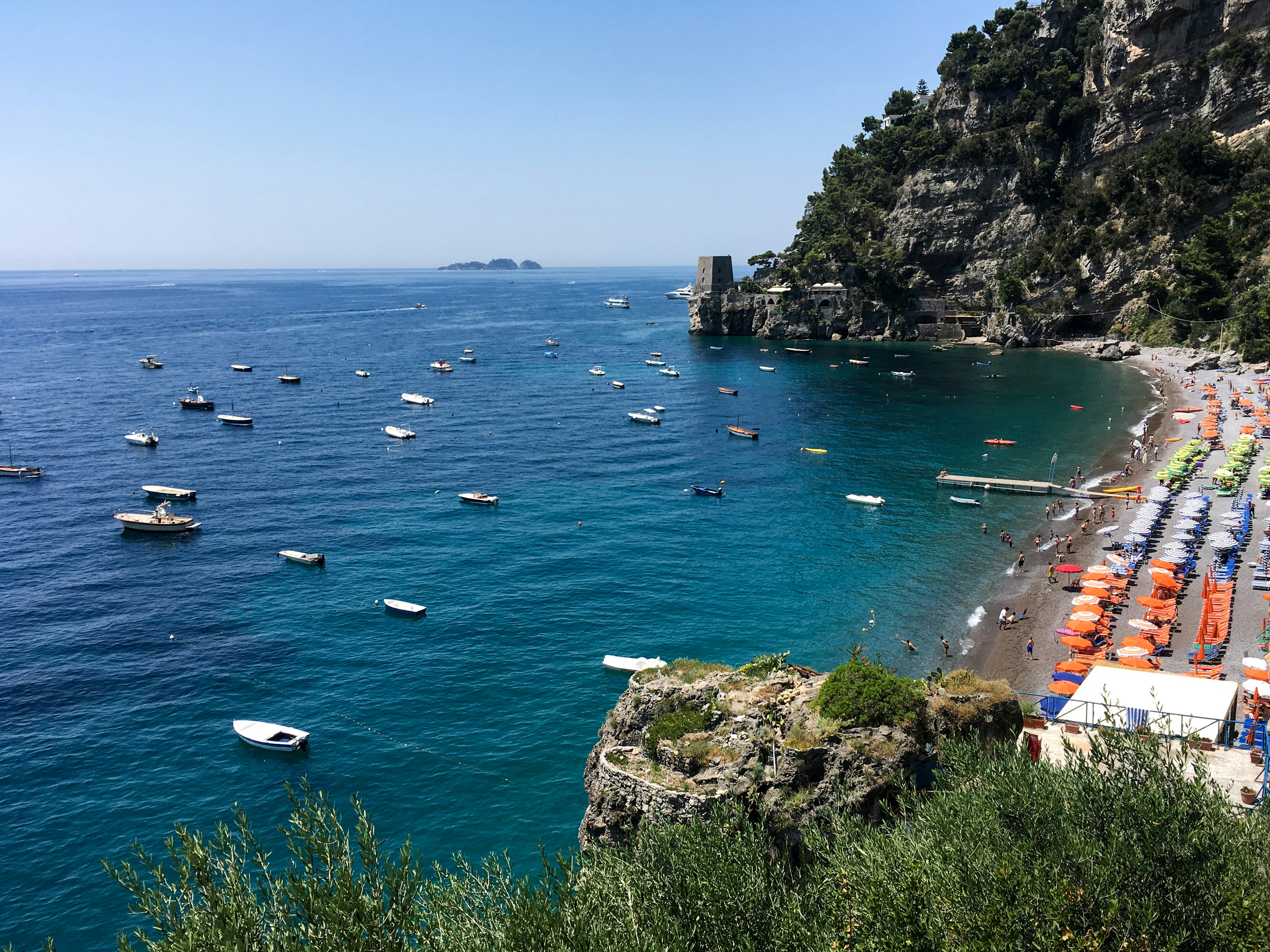 Colorful beach umbrellas line a pebbled shore beside a rocky coastline, with boats dotting the clear blue sea under a sunny sky.