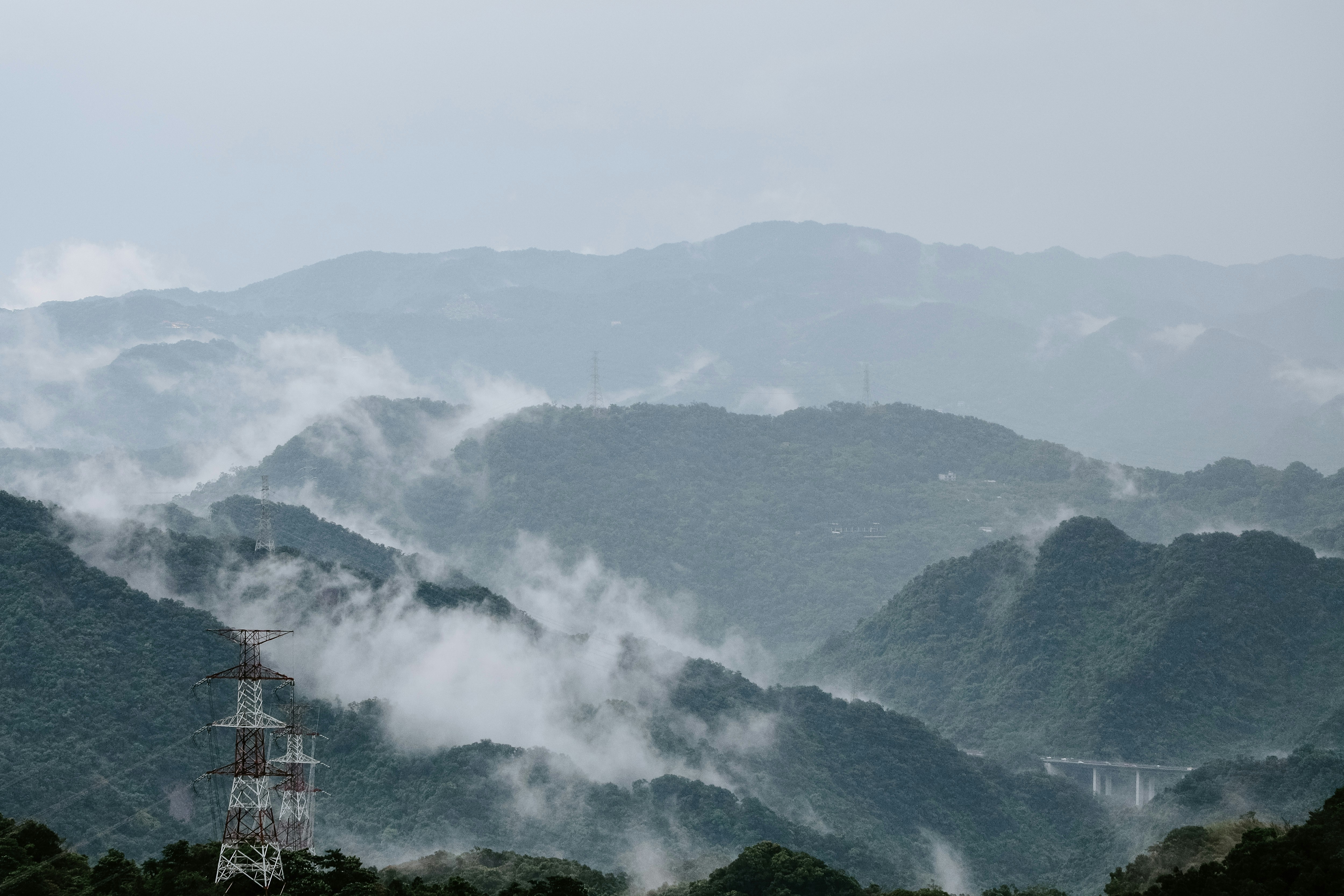 Transmission tower amidst mist-covered mountains under a cloudy sky.