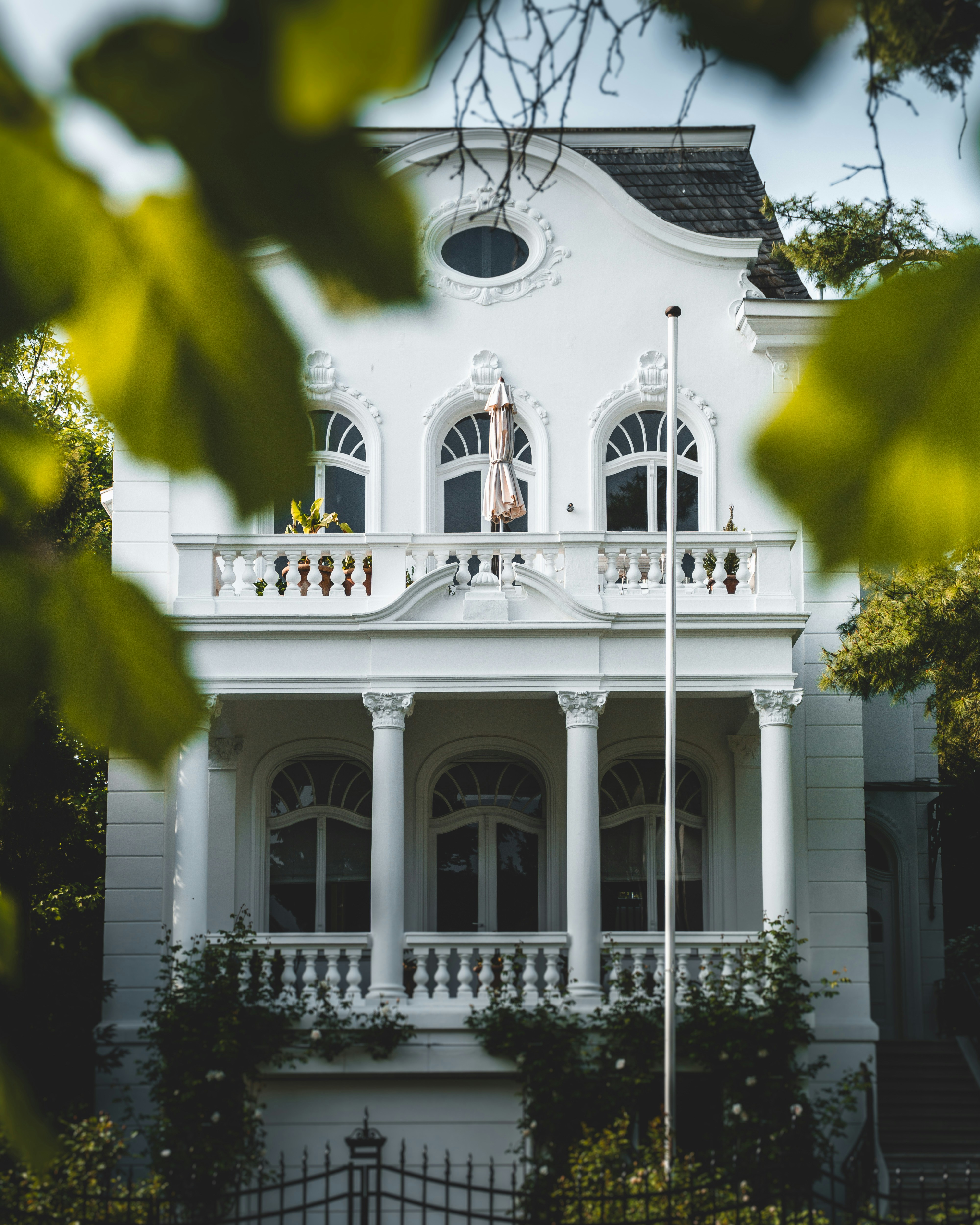 A grand white mansion with intricate architectural details, framed by lush green foliage. A parasol on the balcony adds a touch of charm.