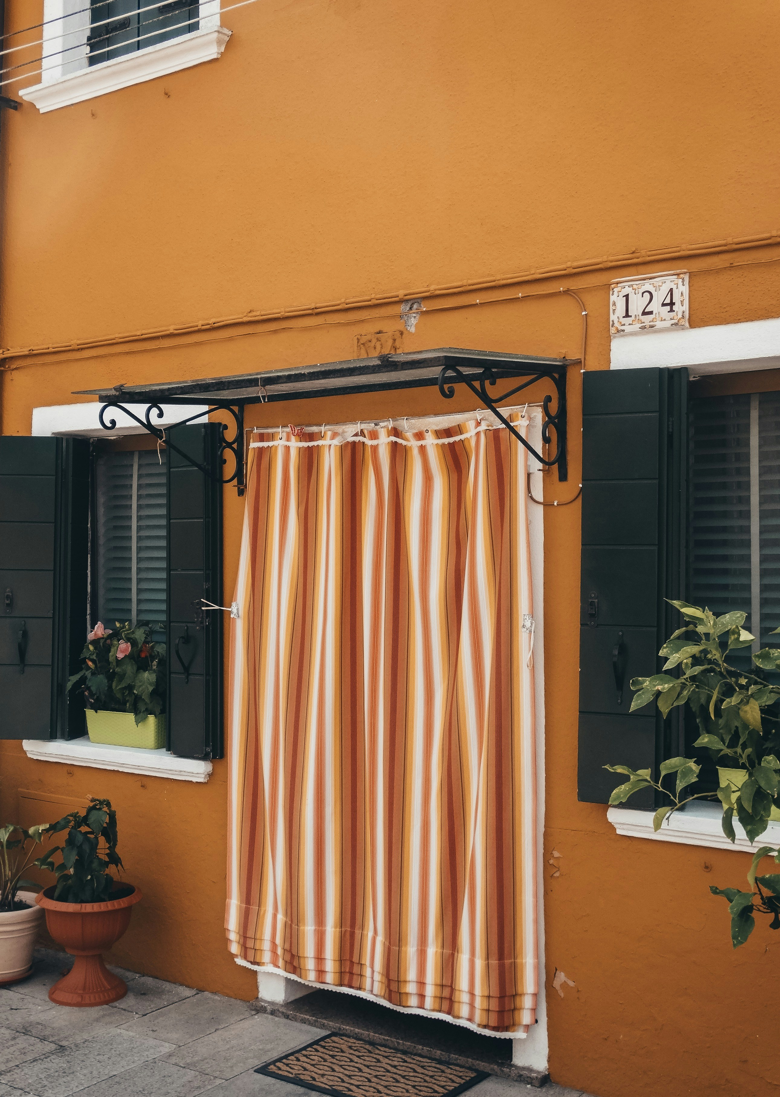 Vibrant striped curtain hangs under an ornate iron awning, framing a charming orange wall with green shutters and potted plants.