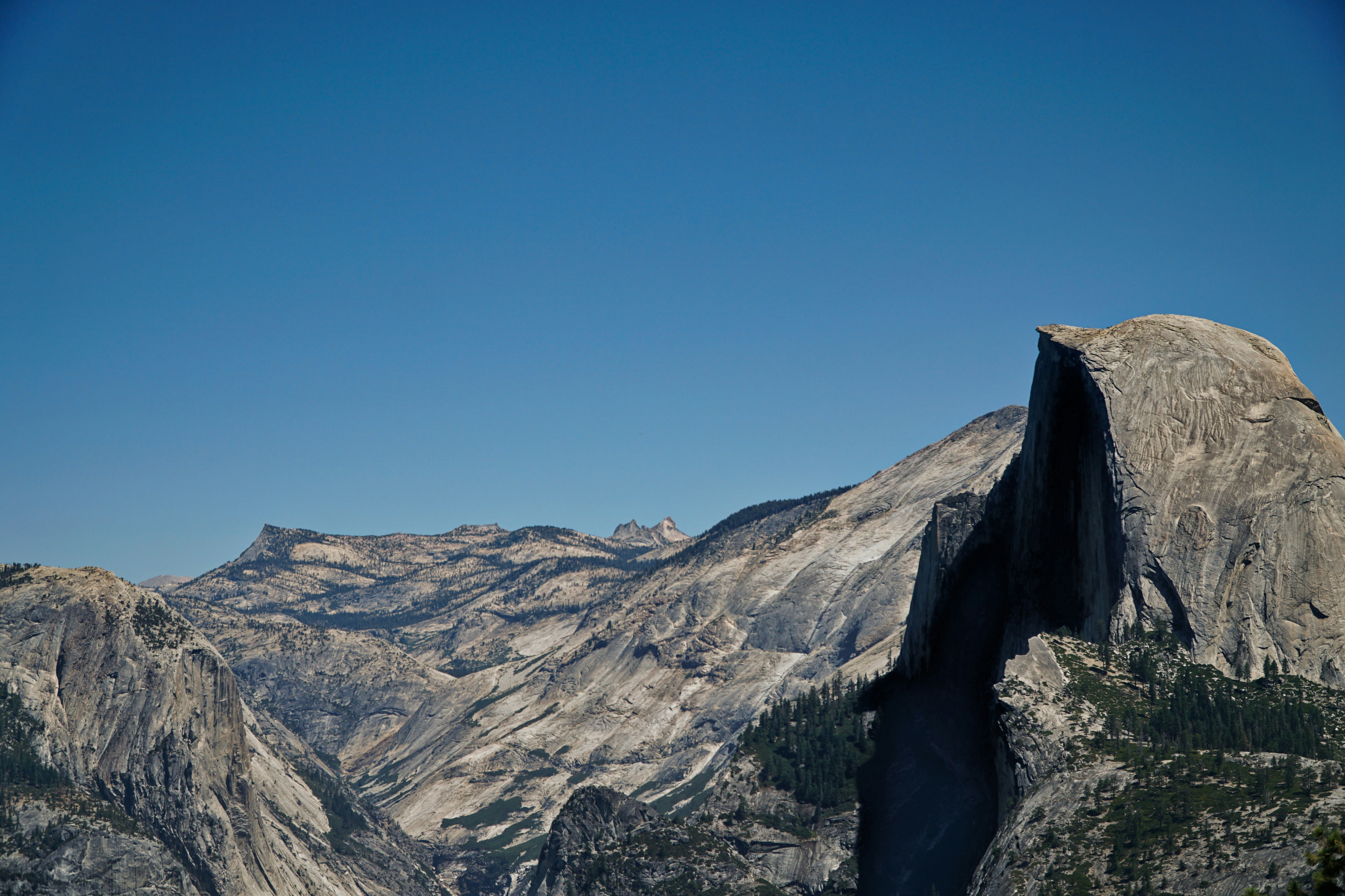 This striking image captures the iconic Half Dome in Yosemite National Park, bathed in natural sunlight against a clear, vibrant blue sky. The composition showcases the rugged granite cliffs and the expansive mountainous landscape, creating a sense of grandeur and timelessness. The contrast between the deep shadows and the sunlit rock formations enhances the dramatic atmosphere, making the scene visually captivating.