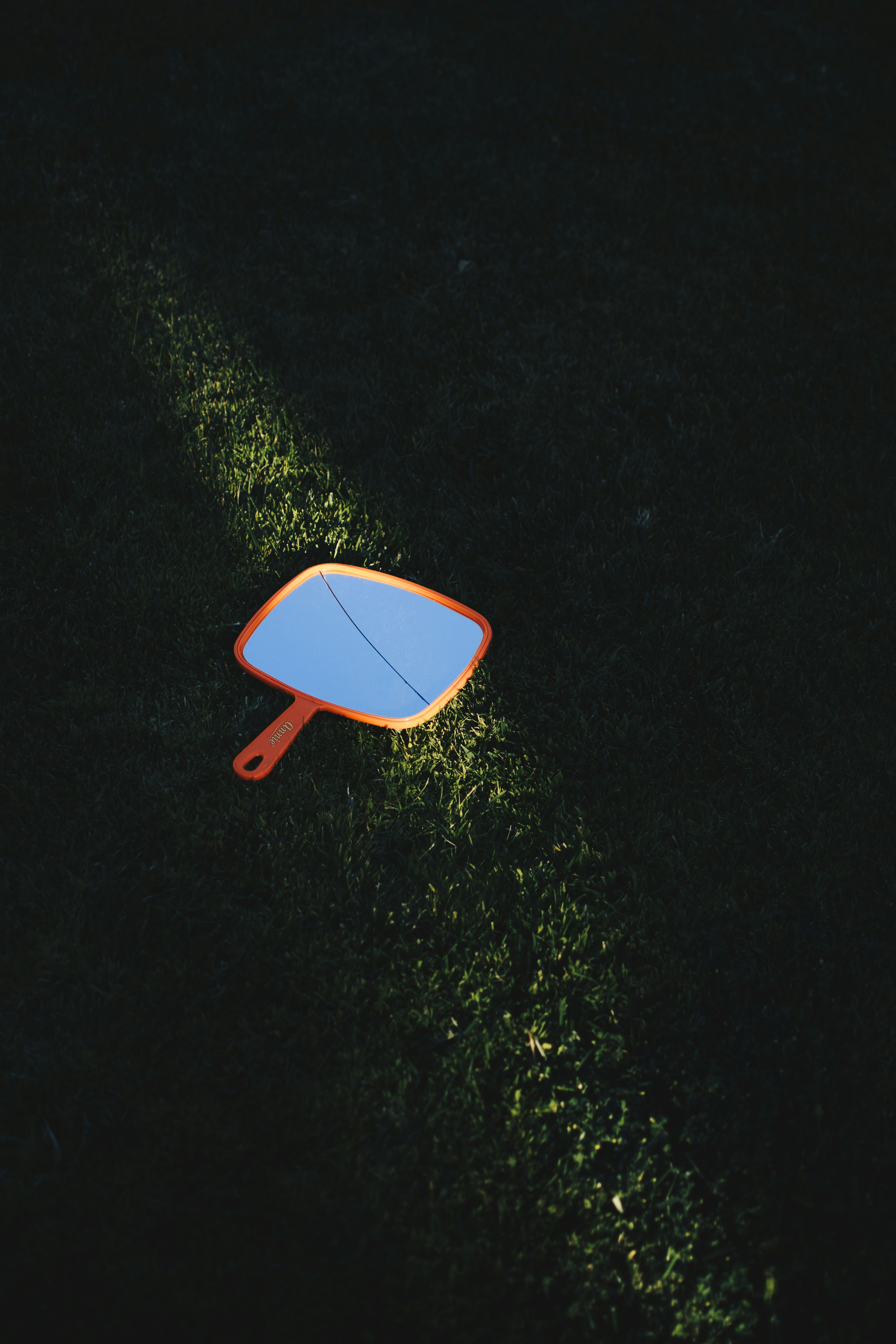 Orange-framed hand mirror resting on a sunlit grass seam