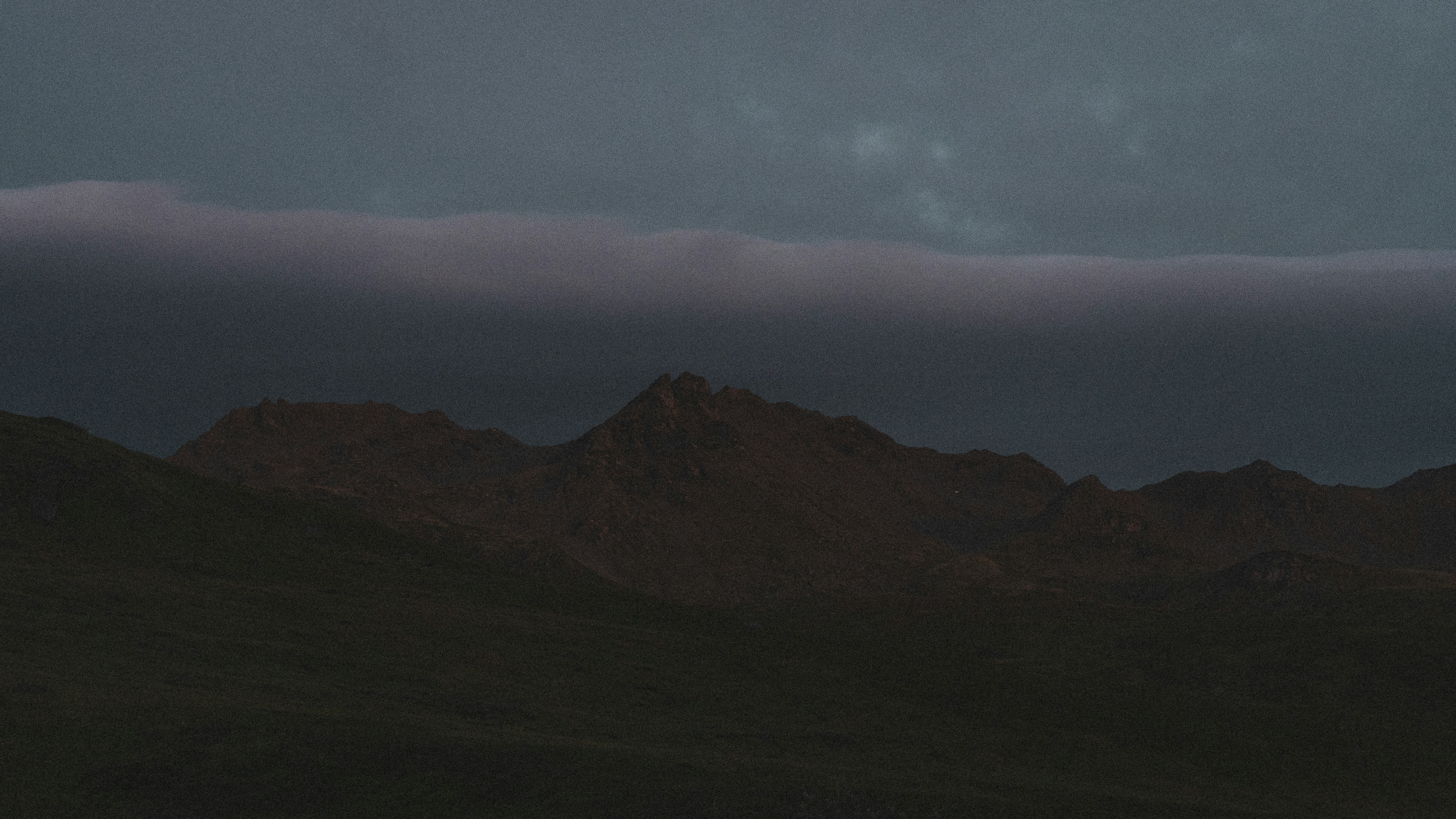 Dark mountain silhouettes emerge beneath a thick layer of clouds, creating a moody atmosphere as twilight descends.
