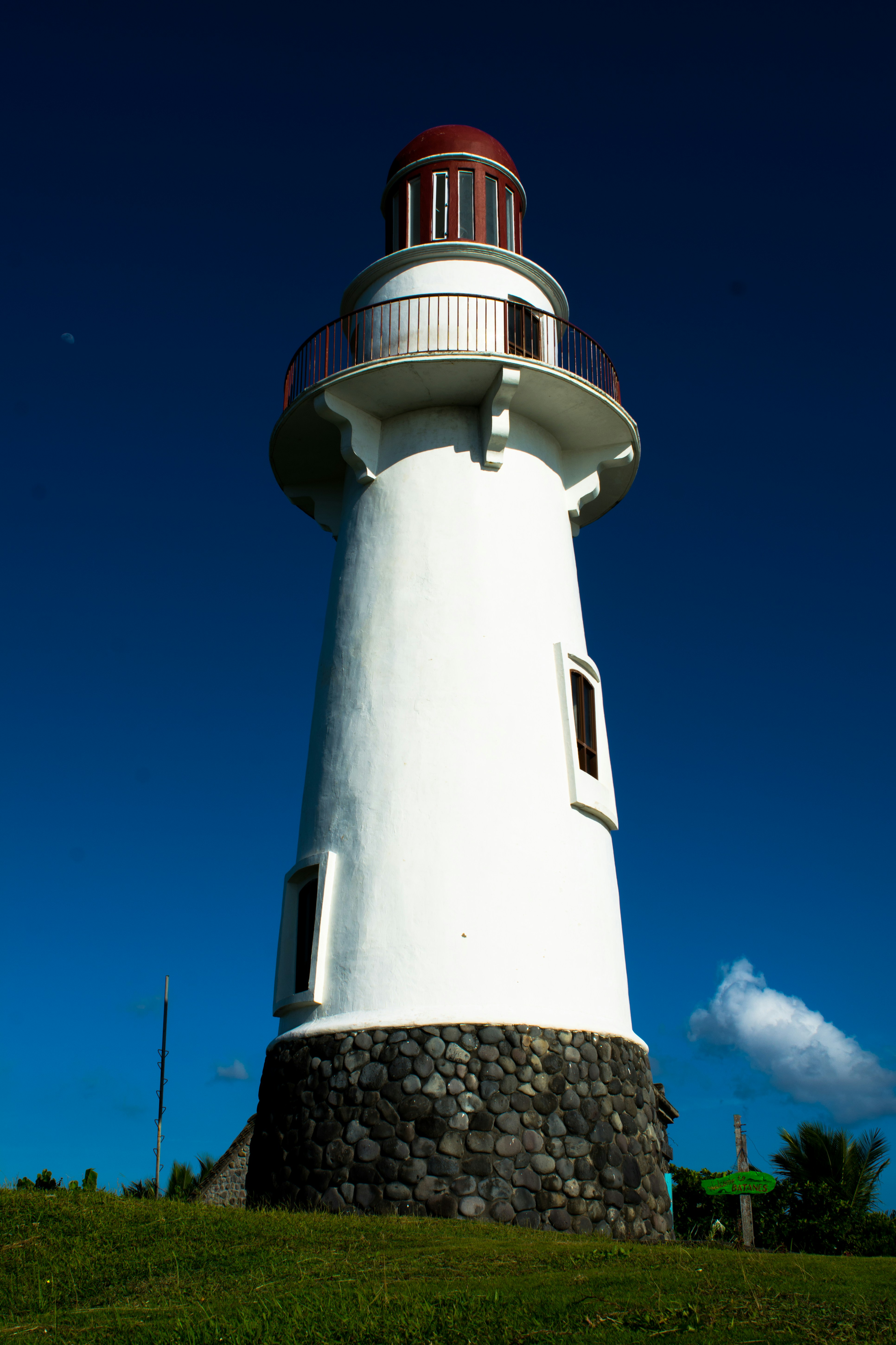A tall lighthouse stands proudly against a clear blue sky, highlighting its white tower and red dome. The structure is surrounded by lush greenery and rocky terrain.