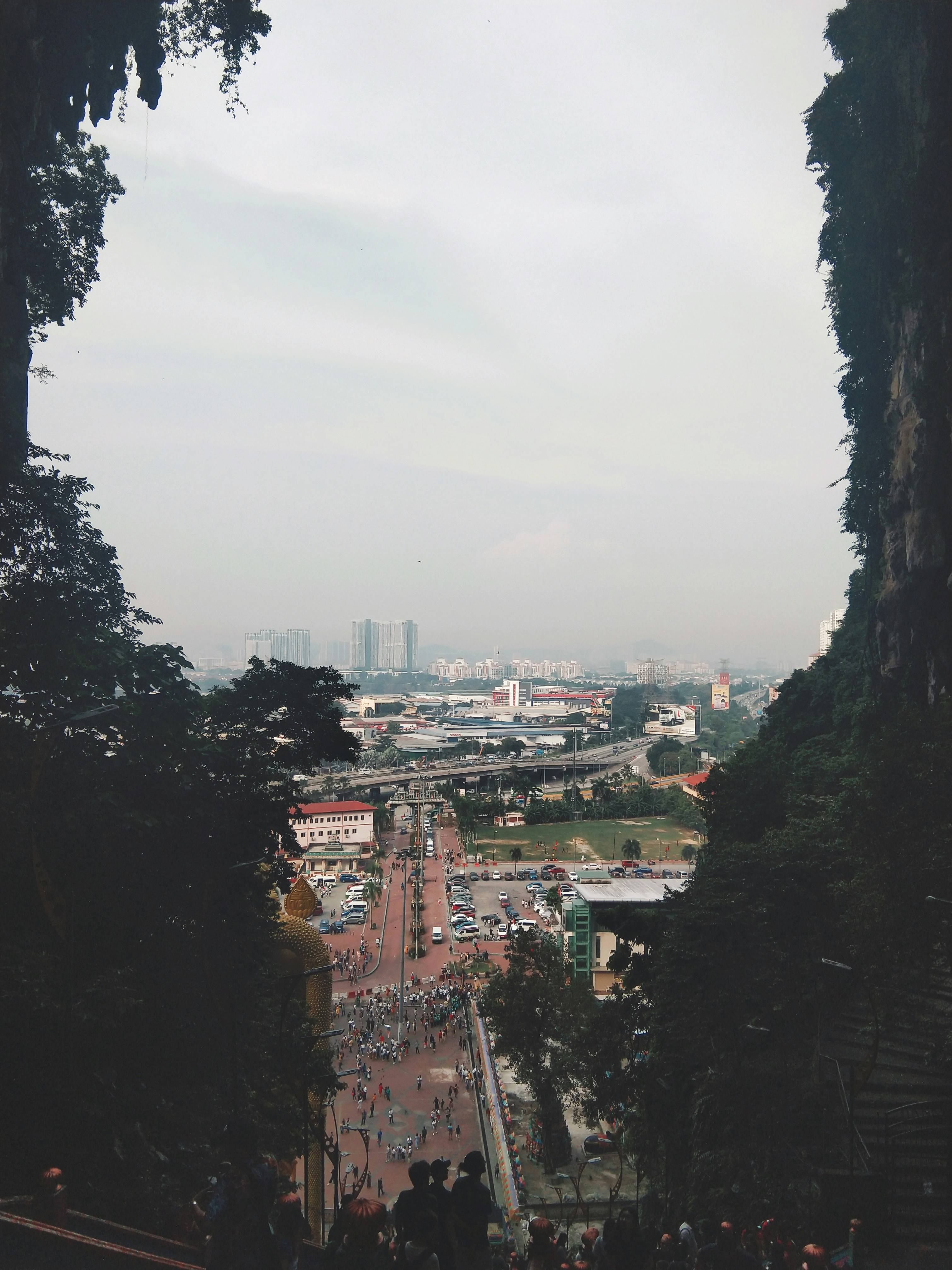 View from a rocky overlook revealing a bustling cityscape below, framed by lush greenery. A pathway leads through the scene, connecting nature with urban life.