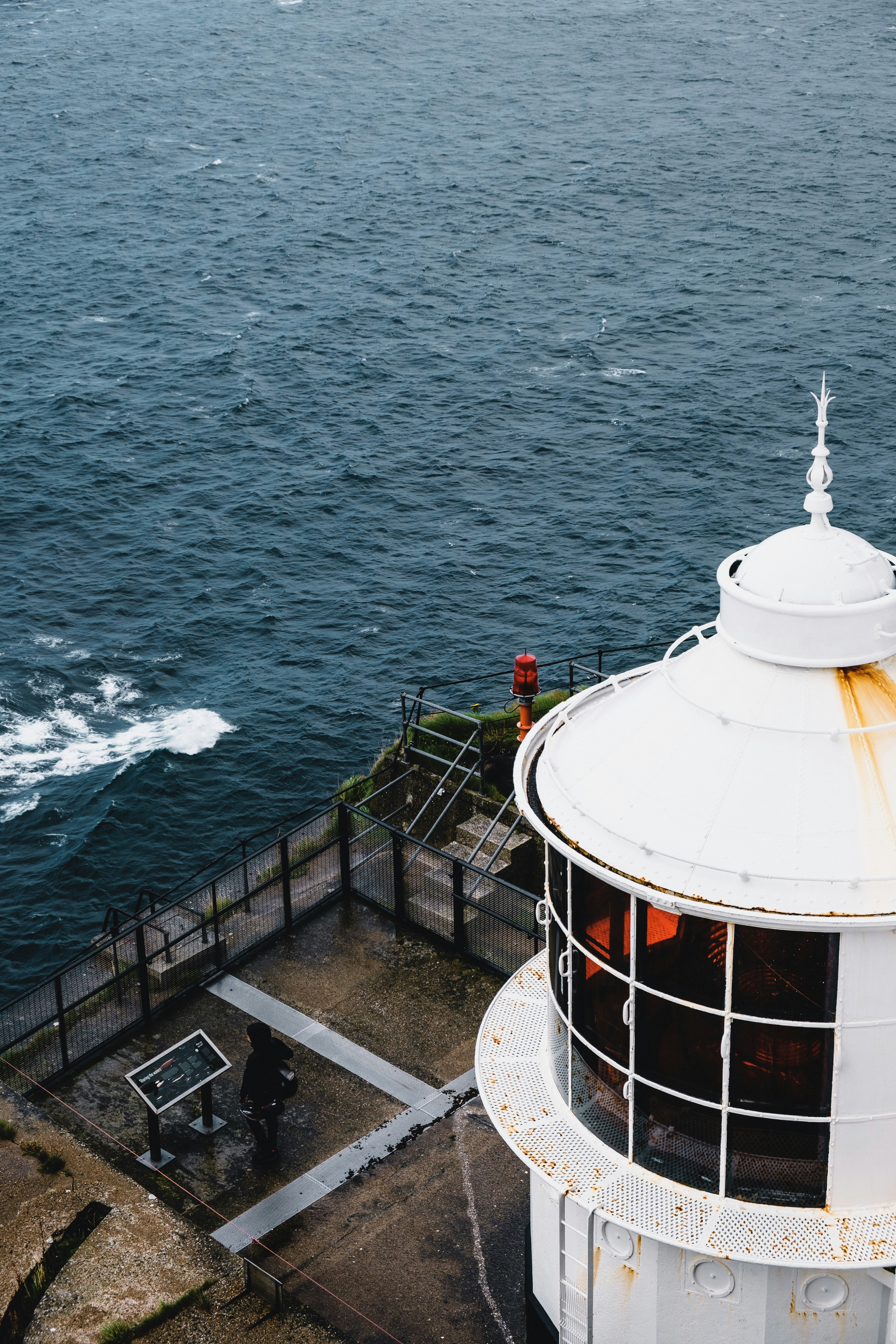 A lighthouse stands sentinel over a turbulent sea, with a visitor observing nearby. The scene captures the interplay of land and ocean.