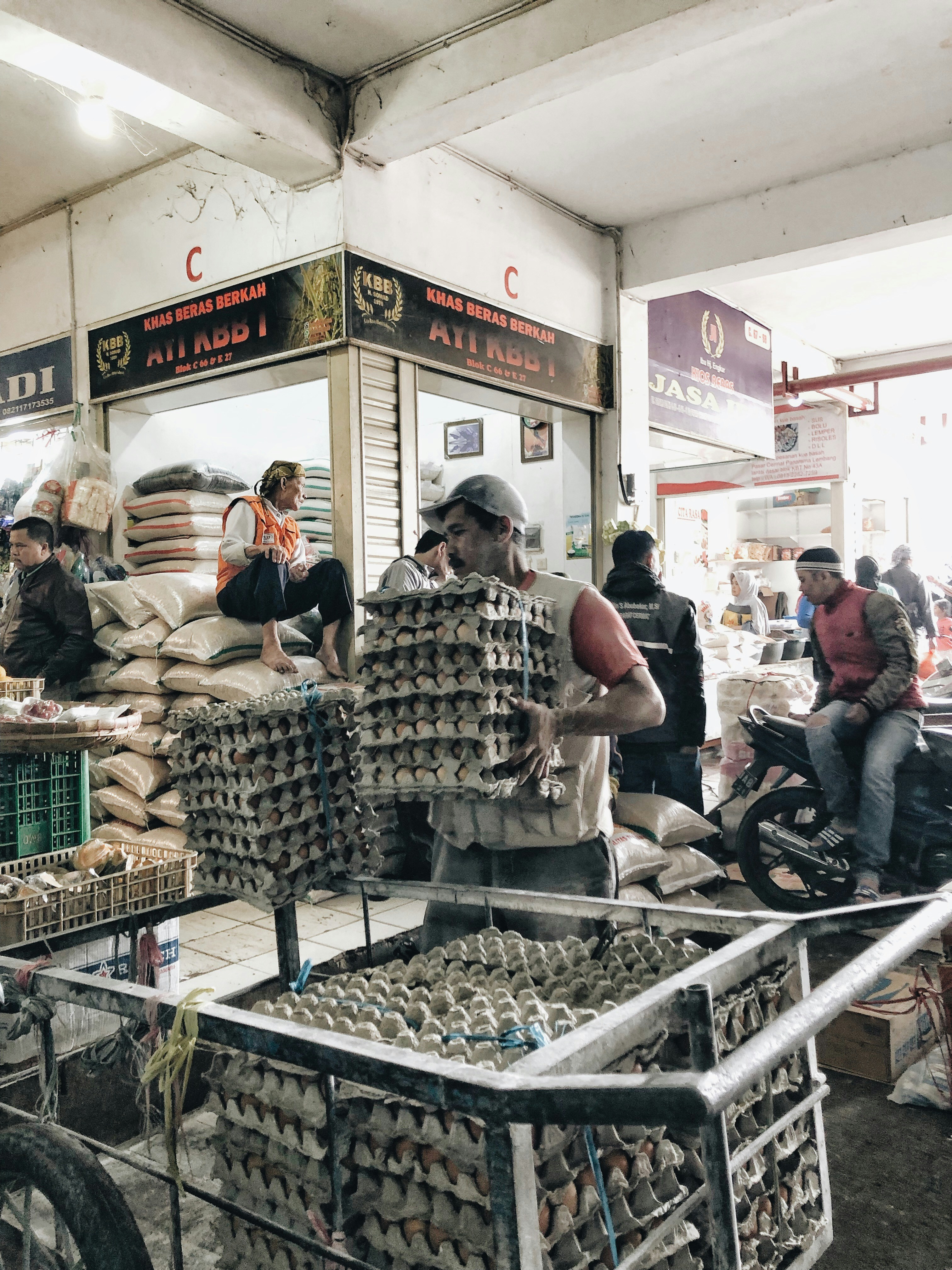 Vendor carrying stacks of eggs through a busy indoor market with bags of rice and patrons in the background.