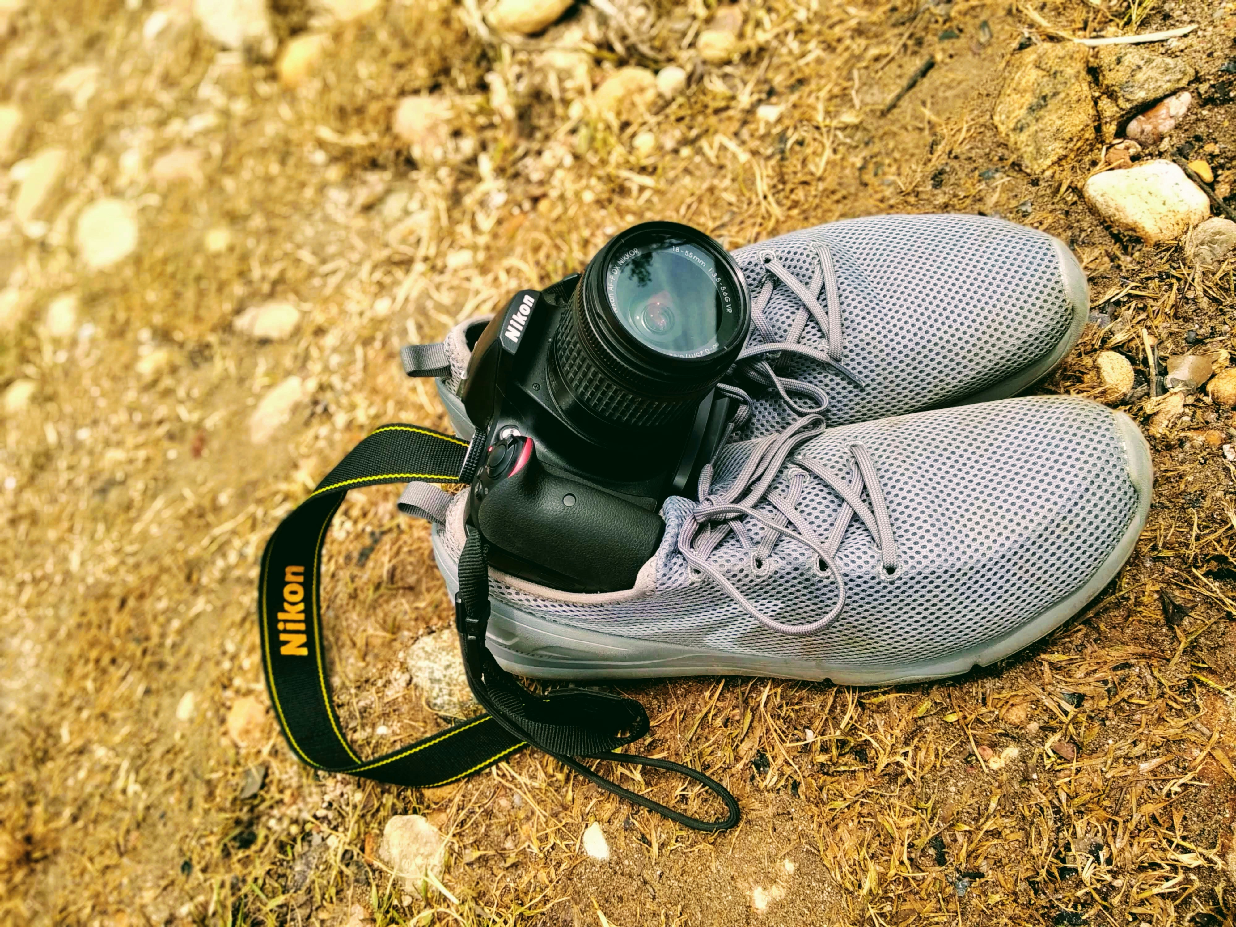 A pair of grey athletic shoes with a Nikon camera resting on top, set against a natural, earthy background. The scene conveys a sense of readiness for outdoor photography.