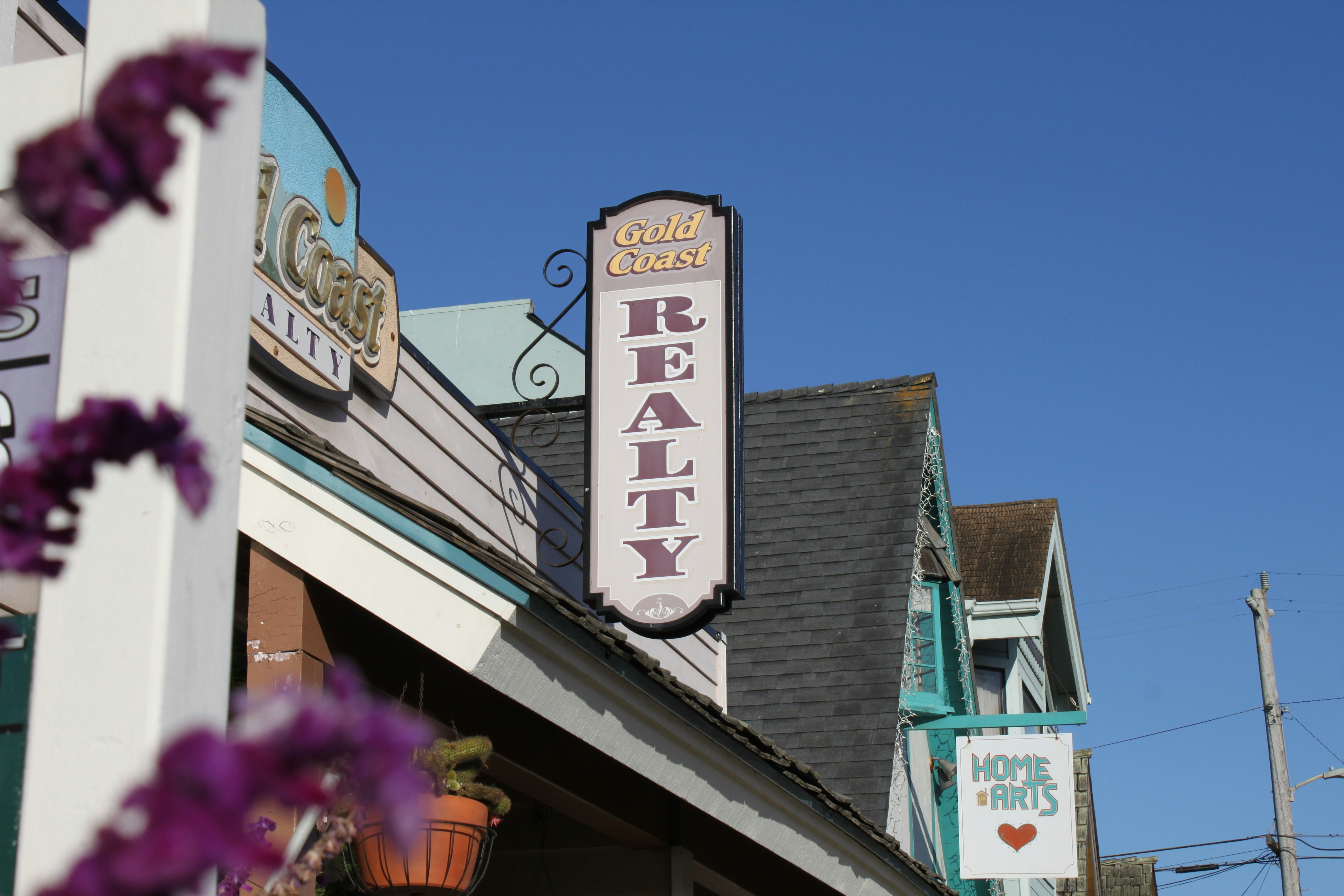 Storefront photograph featuring a tall 'Gold Coast Realty' sign and a vertical 'REALTY' banner against a clear blue sky. Purple flowers frame the foreground.