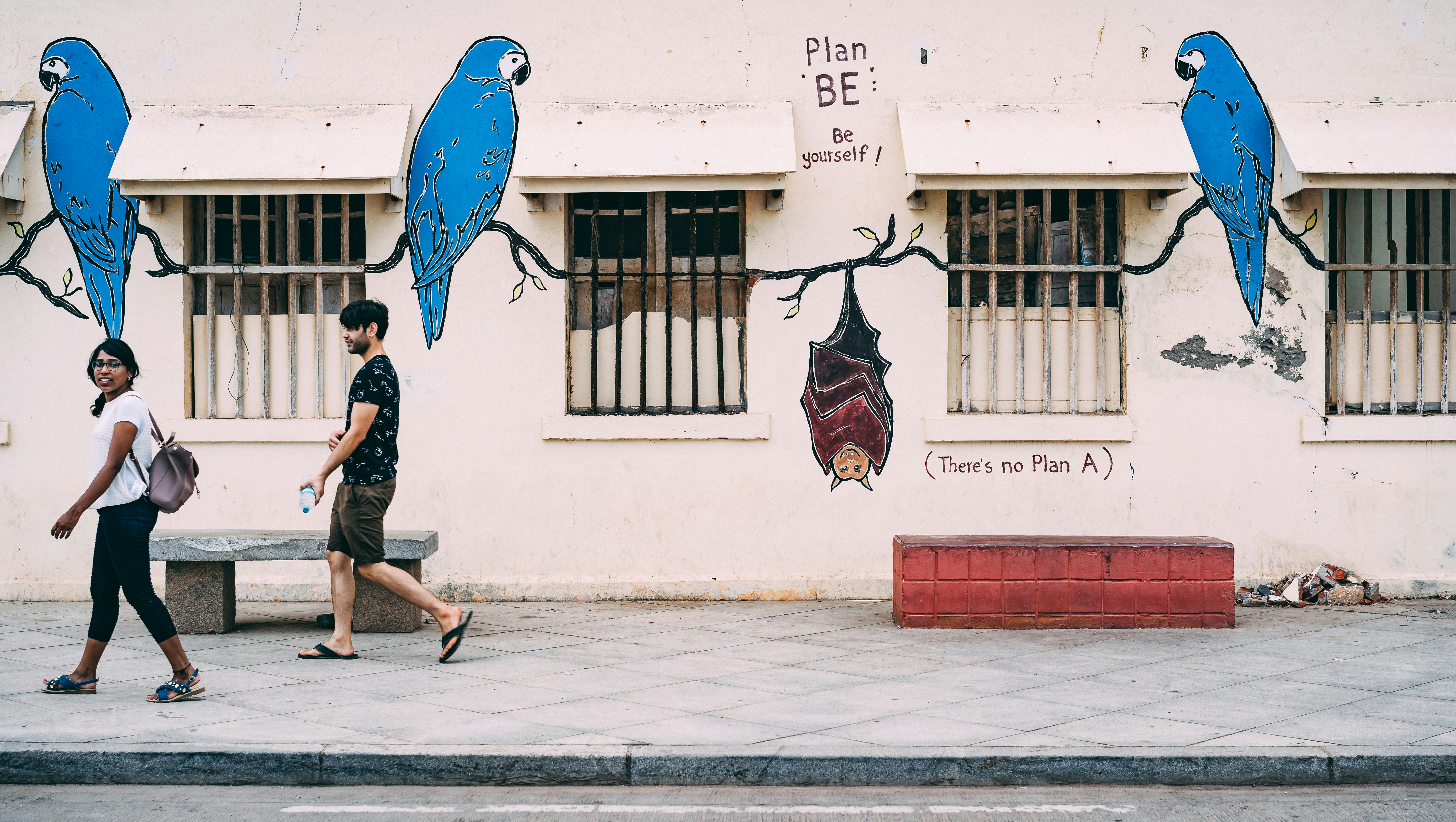 Two pedestrians stroll past a vibrant mural featuring blue parrots and a whimsical bat, blending urban life with artistic expression.