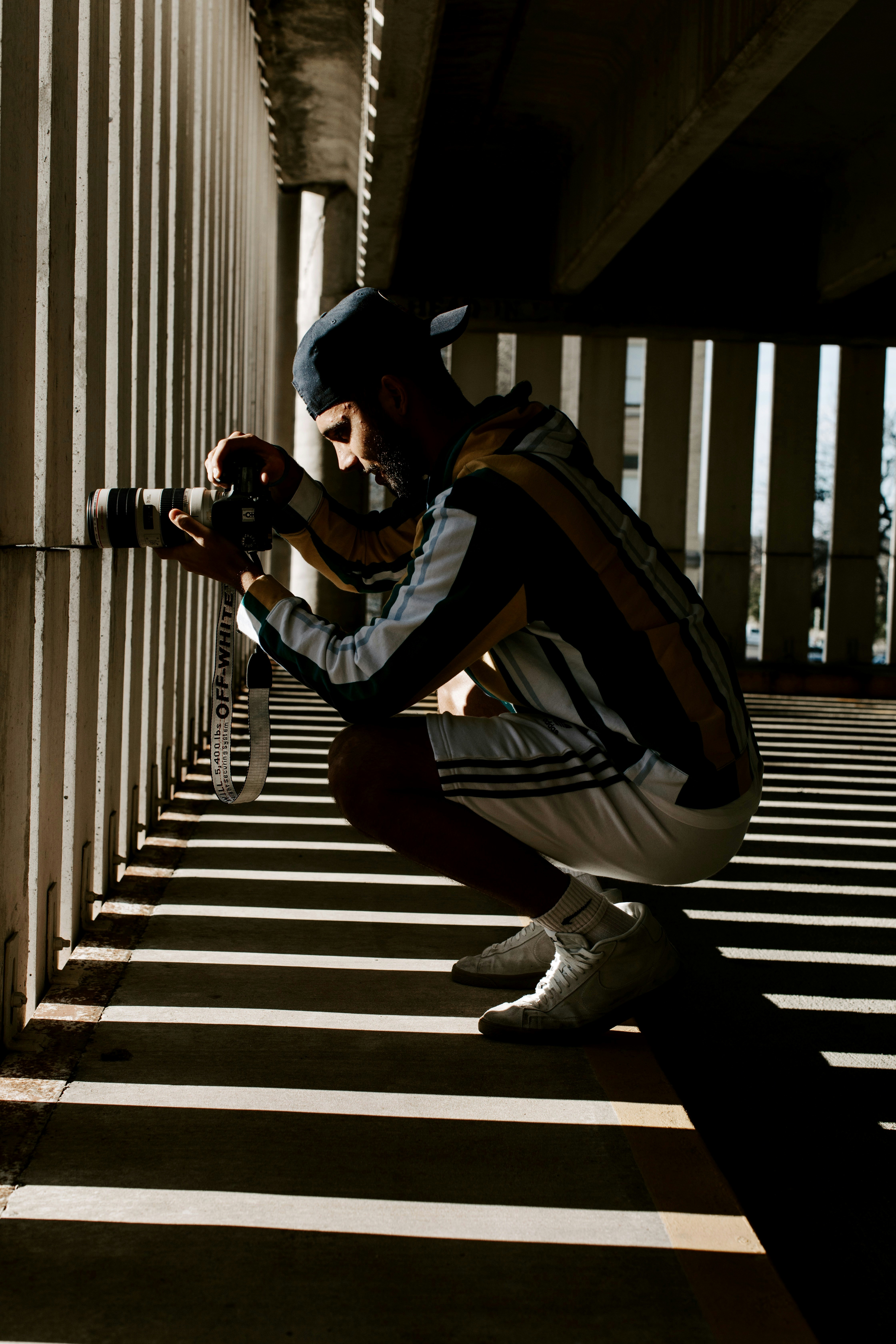 A photographer squatting in a striped shadow pattern while capturing a moment with a Canon camera. The interplay of light and shadow creates a dynamic urban atmosphere.