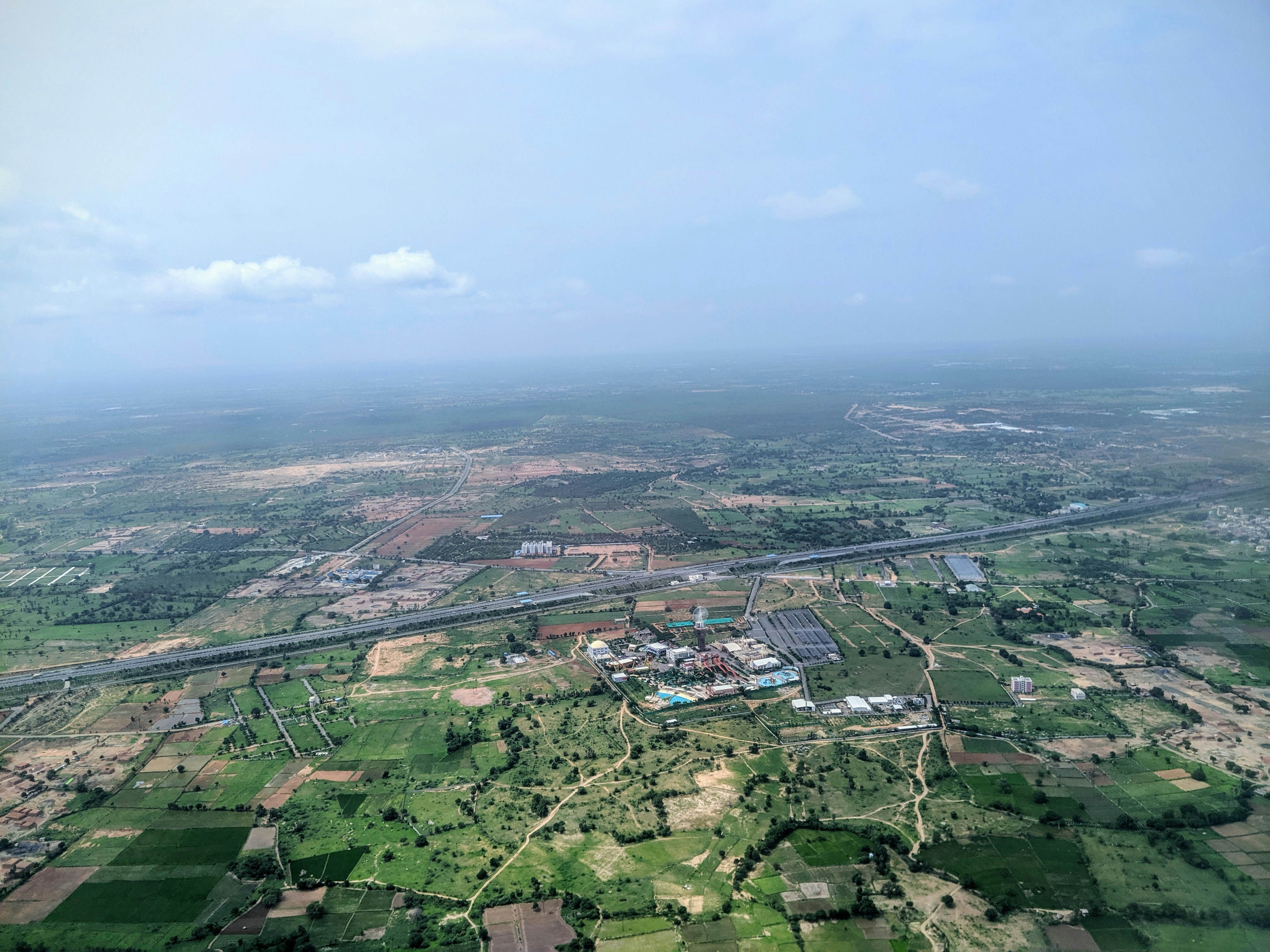 Aerial view showcasing a patchwork of fields and structures, with a highway weaving through the landscape. The scene is enveloped in a soft, cloudy sky.