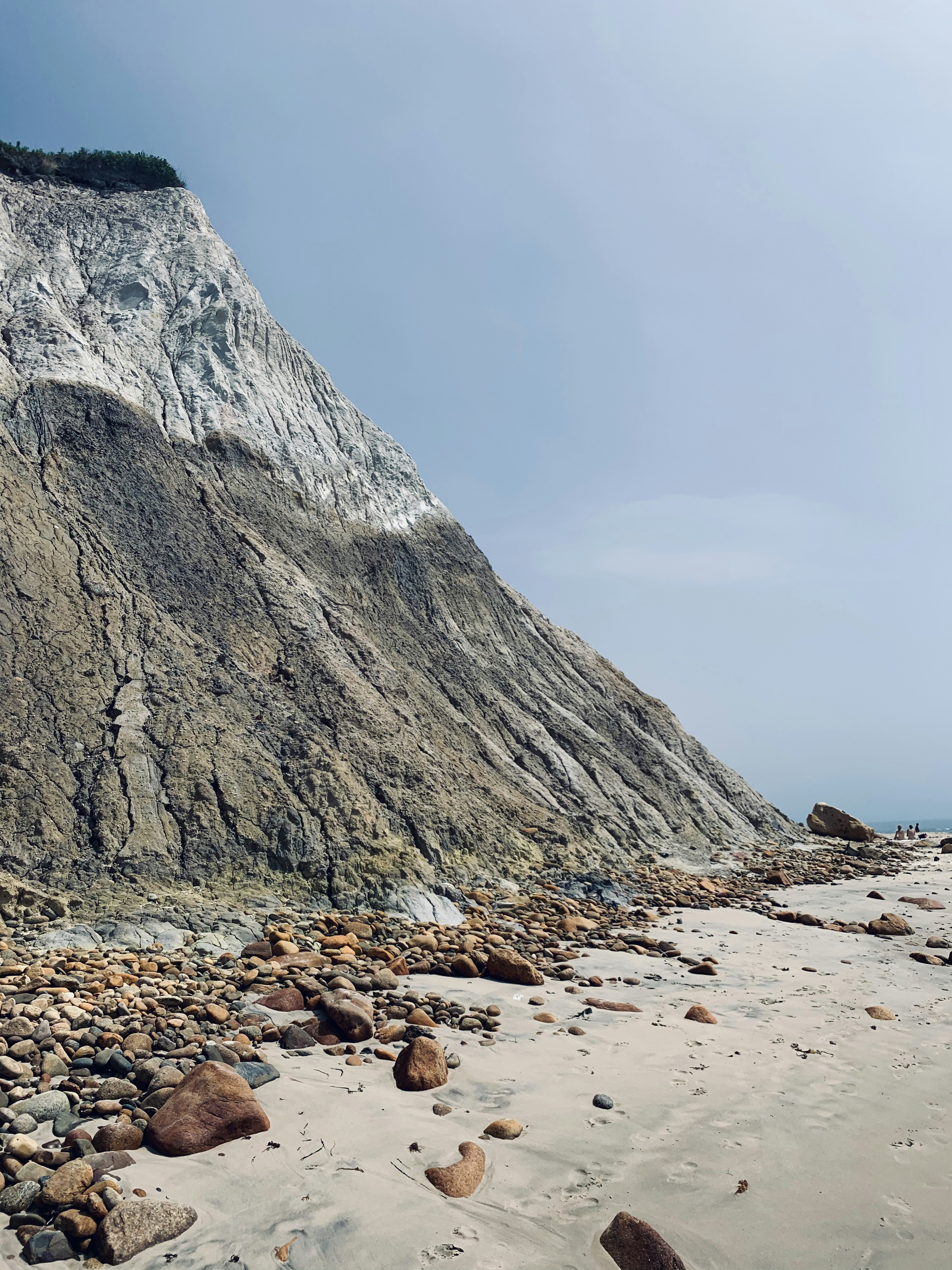 Majestic coastal cliffs rise dramatically above a sandy beach scattered with smooth stones. The scene captures the interplay of earth and sea under a soft, hazy sky.