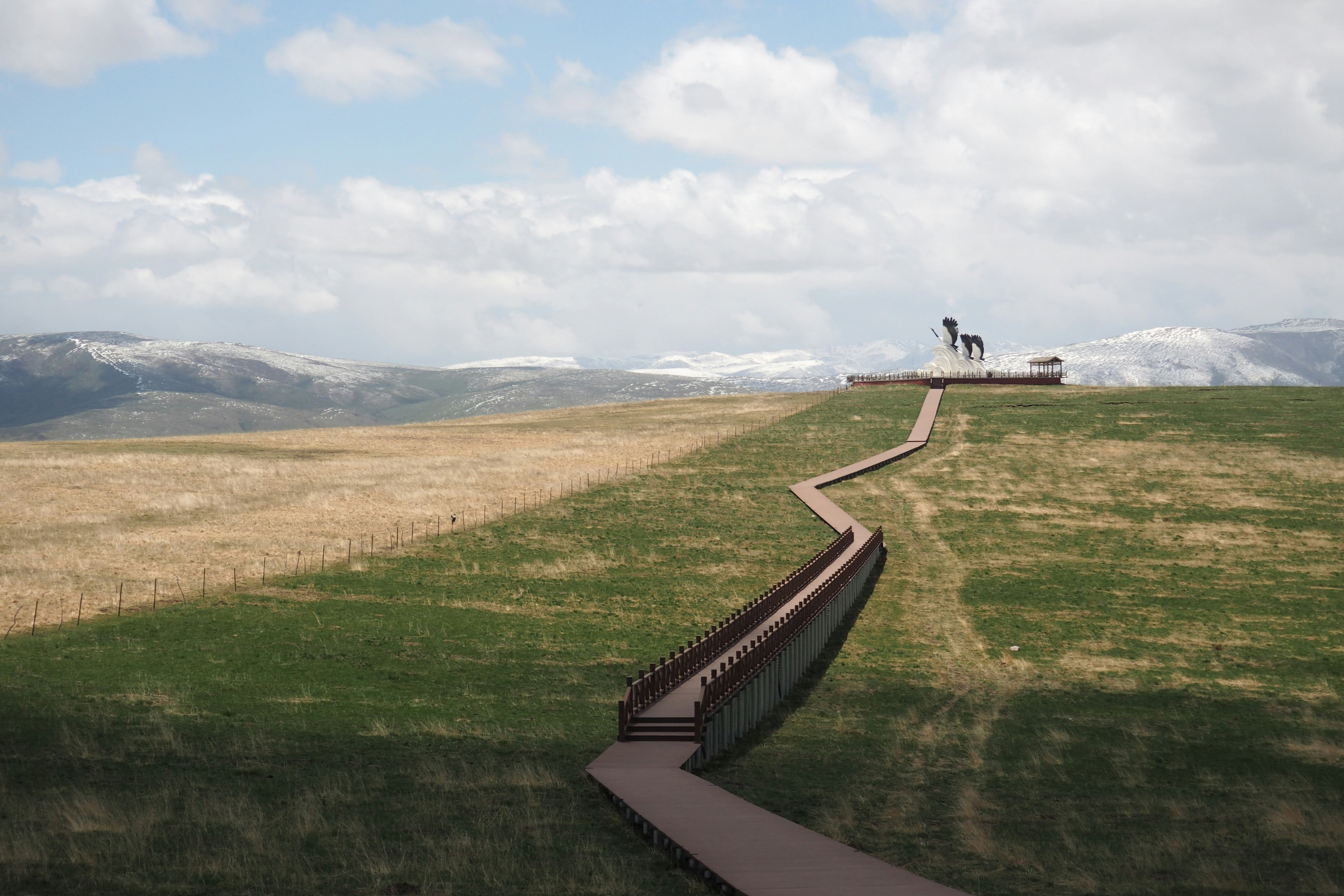 Wooden walkway leading through grassland towards a distant shrine with mountains in the background.
