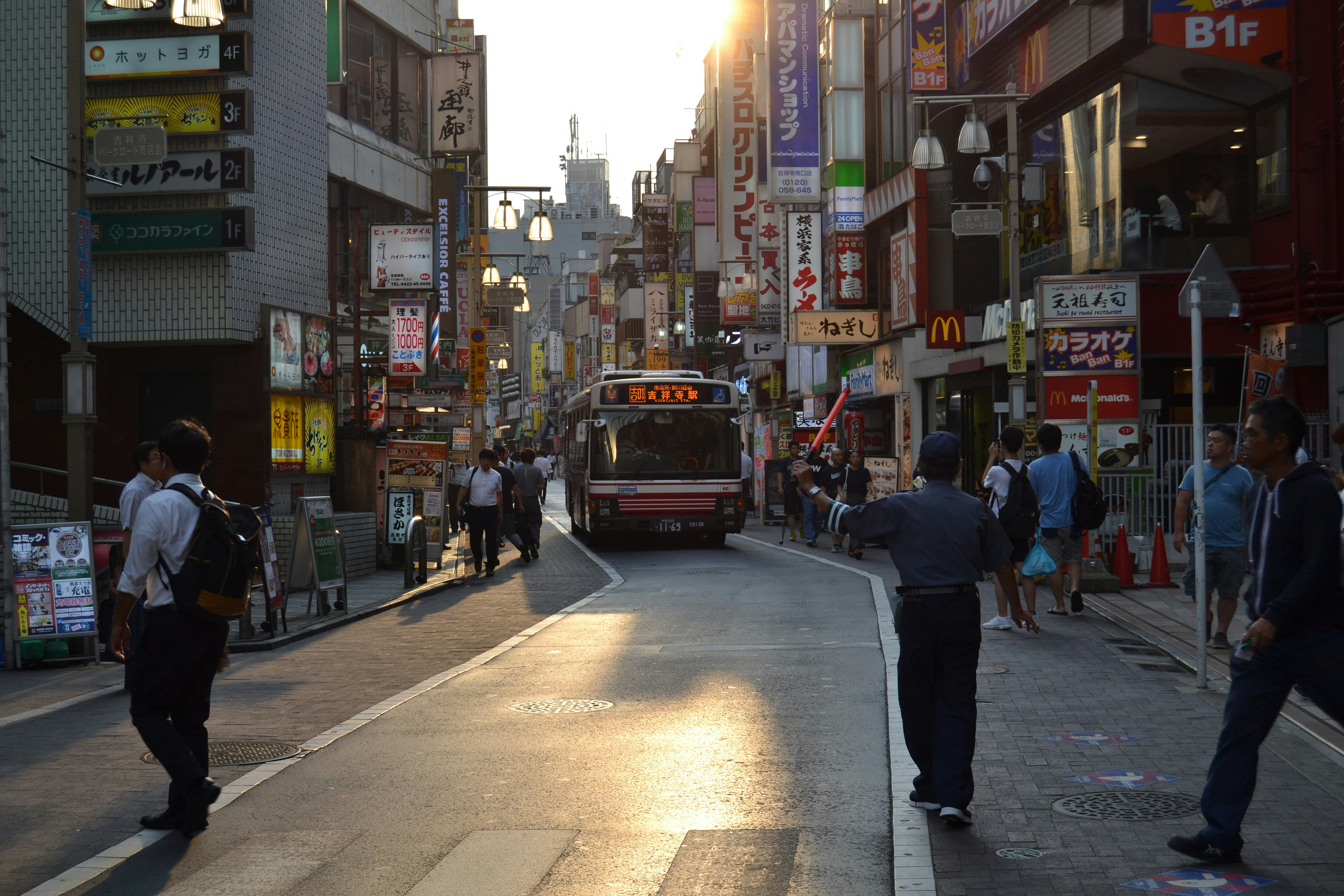 Pedestrians and a bus navigate a narrow city street under the warm glow of the setting sun.