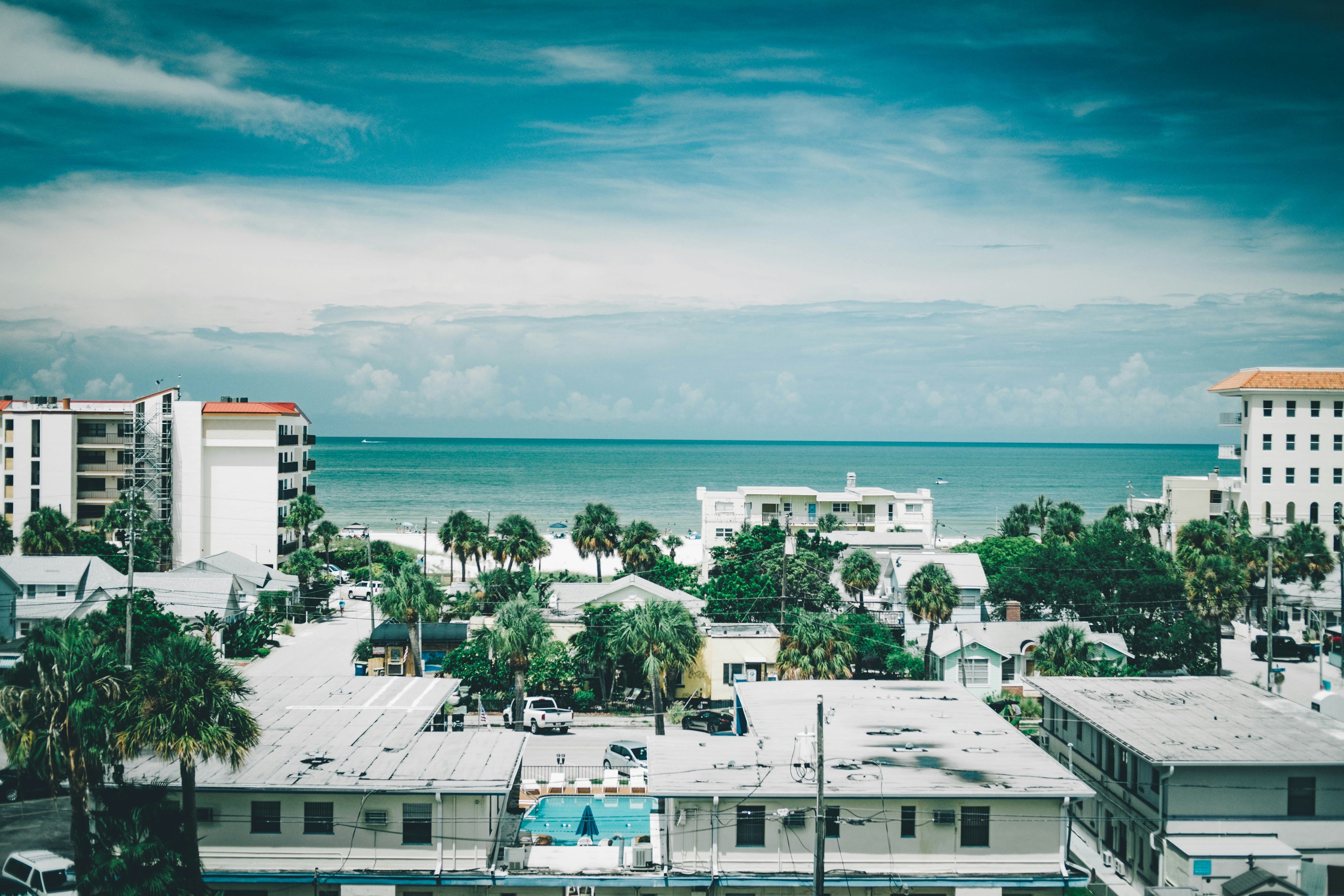 A panoramic view of a coastal town with residential buildings and palm trees, leading to a serene beach and ocean under a cloudy sky.