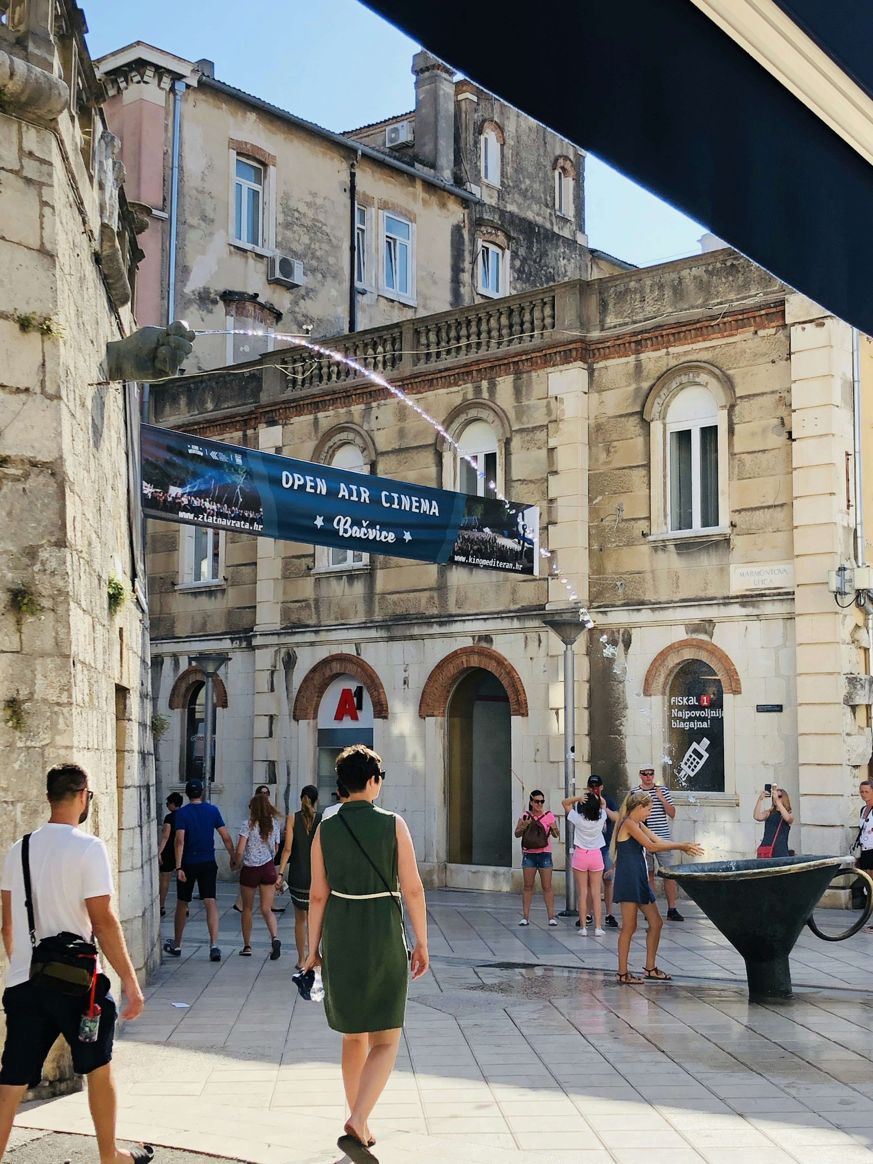 Green-dressed pedestrian under Open Air Cinema banner in a sunlit historic plaza