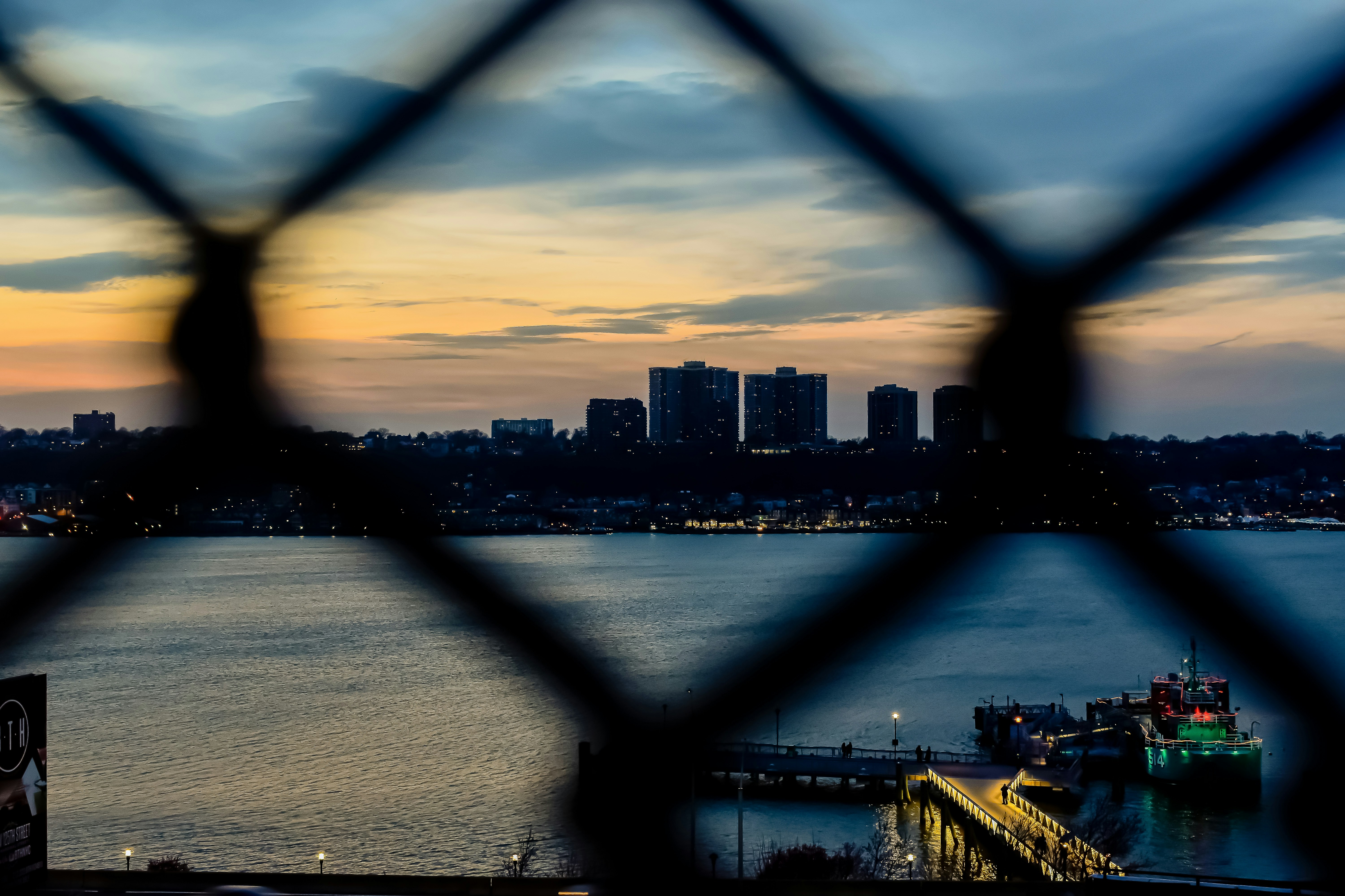 City skyline viewed through a chain link fence at dusk, with shimmering water and an illuminated pier.