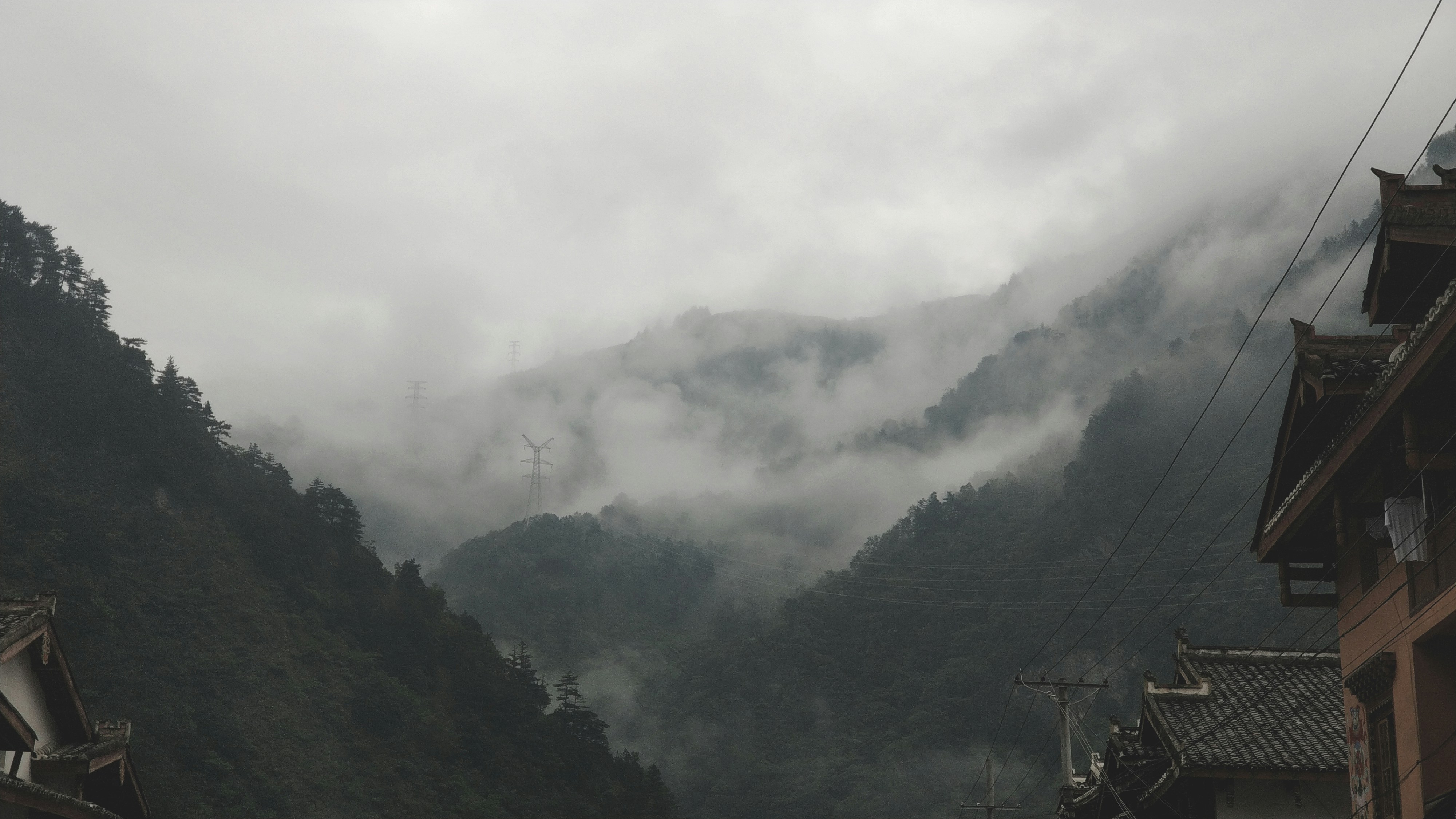 Fog drapes over lush green mountains, with traditional rooftops in the foreground under an overcast sky.