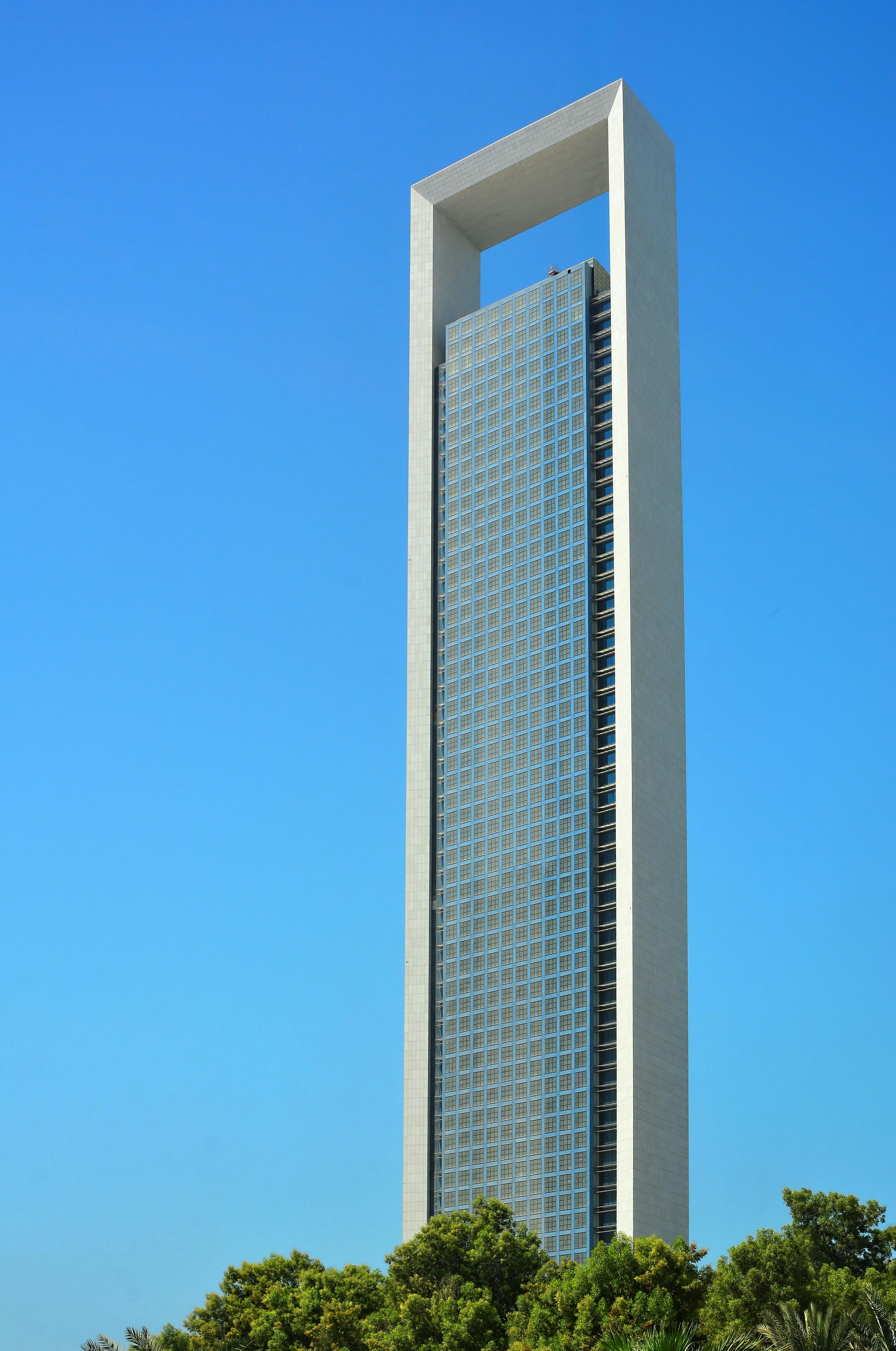 A modern skyscraper featuring a unique open-top design against a clear blue sky, surrounded by lush greenery.