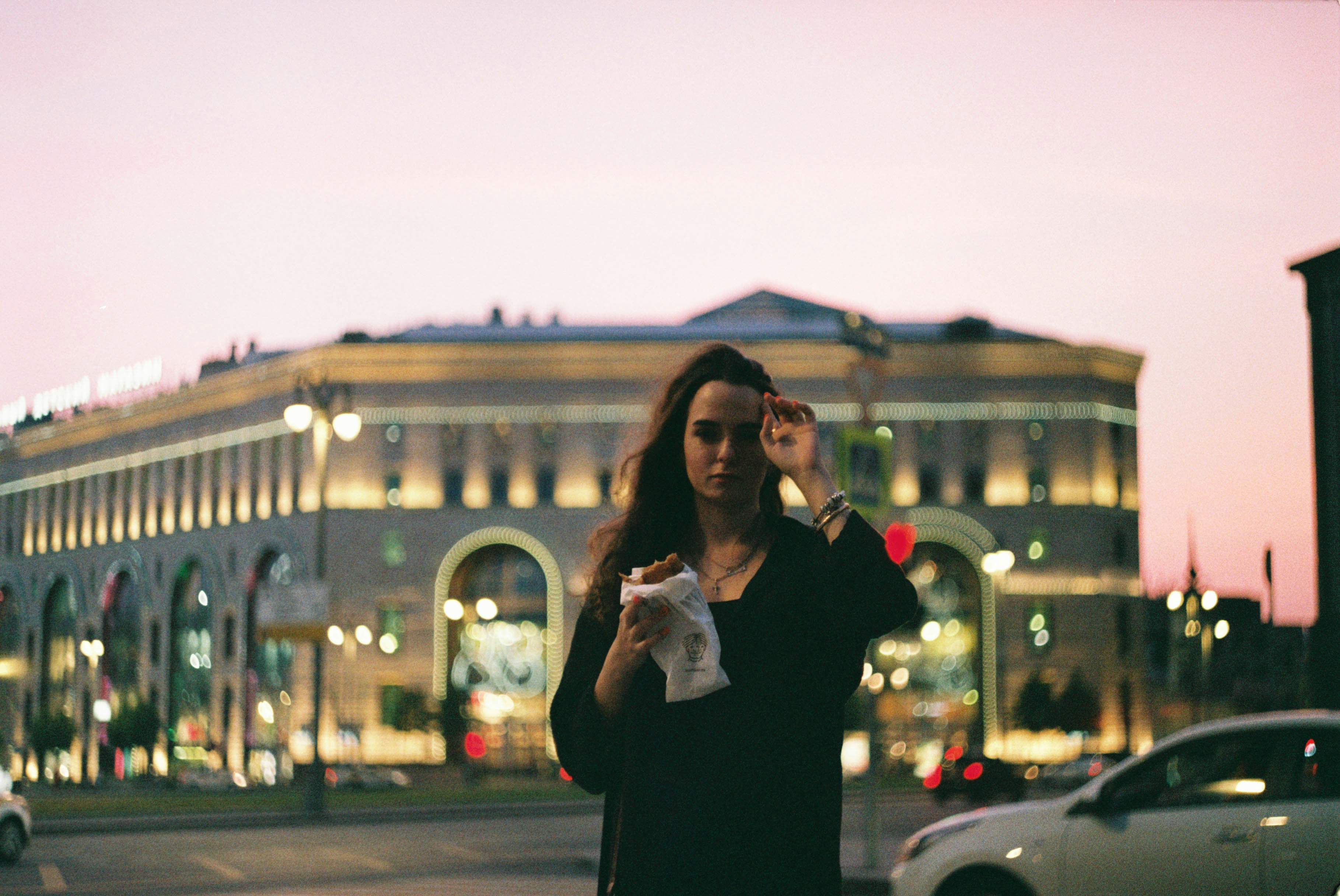 Portrait photograph of a woman in a dark jacket holding a tissue, standing before a softly blurred cityscape at pink twilight.