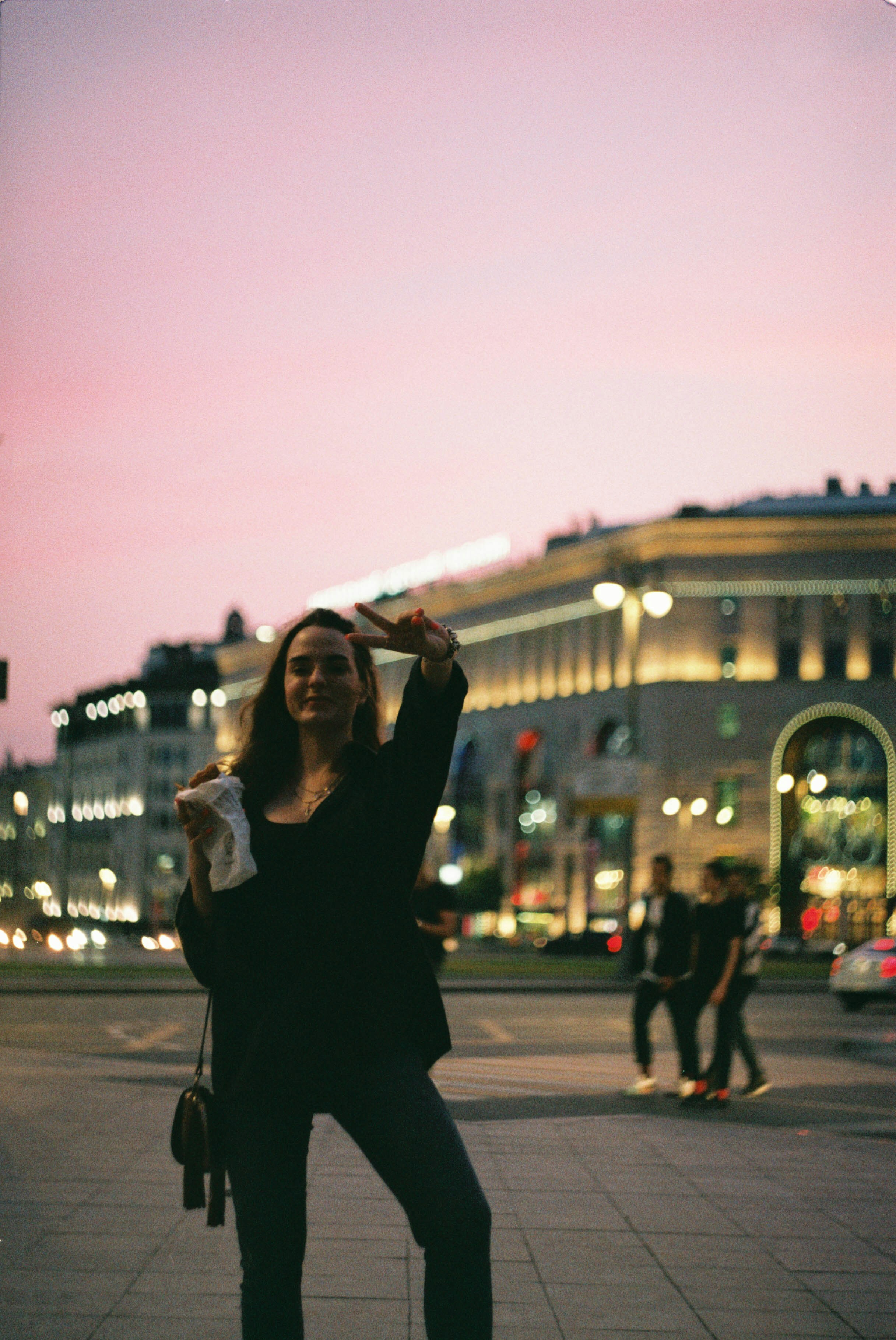 Young woman holding a water bottle and making a peace sign against a vibrant twilight sky, with city lights softly illuminating the background.