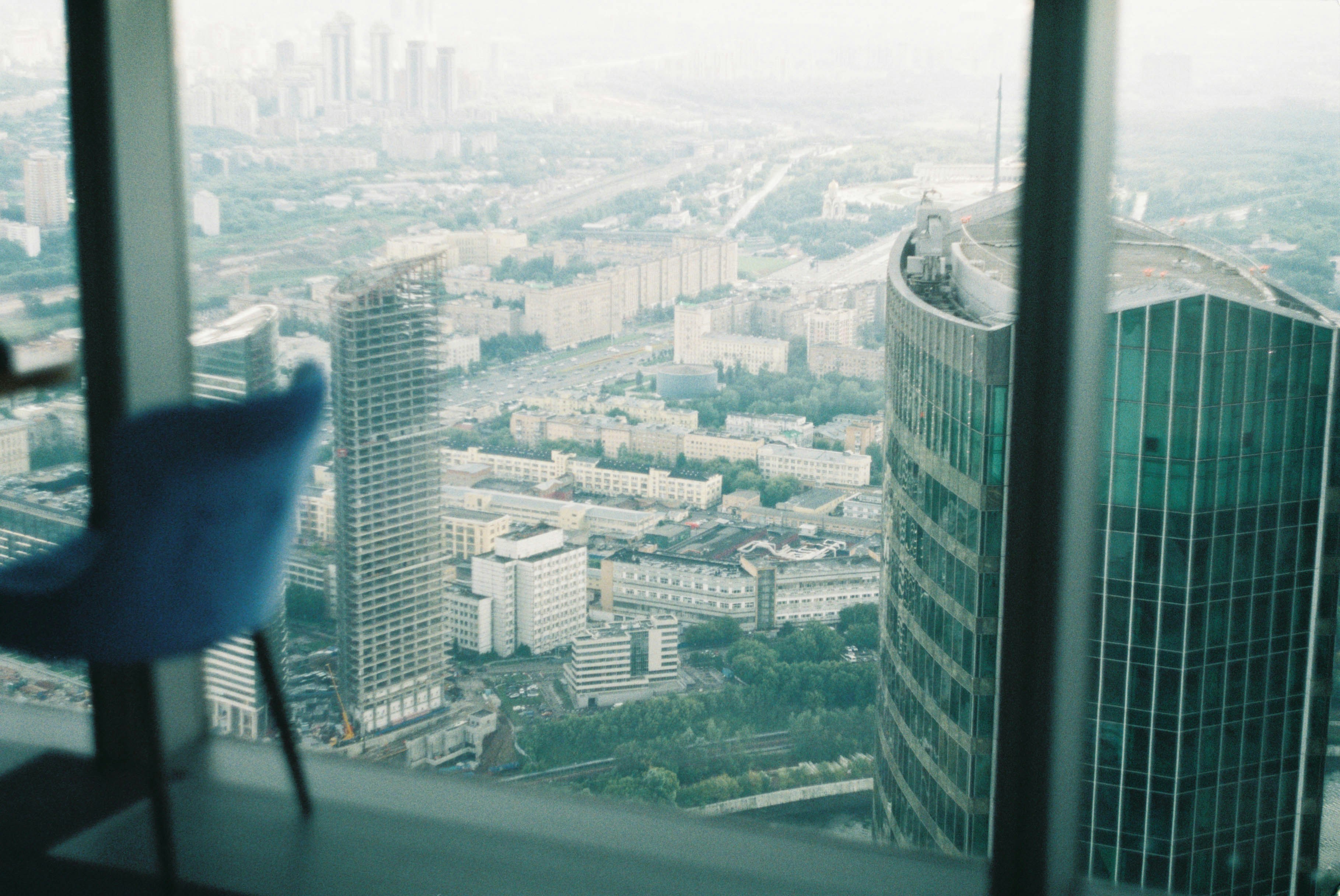 A panoramic view of a bustling cityscape featuring modern skyscrapers and a winding road seen through a glass window. The composition includes a stylish blue chair in the foreground.
