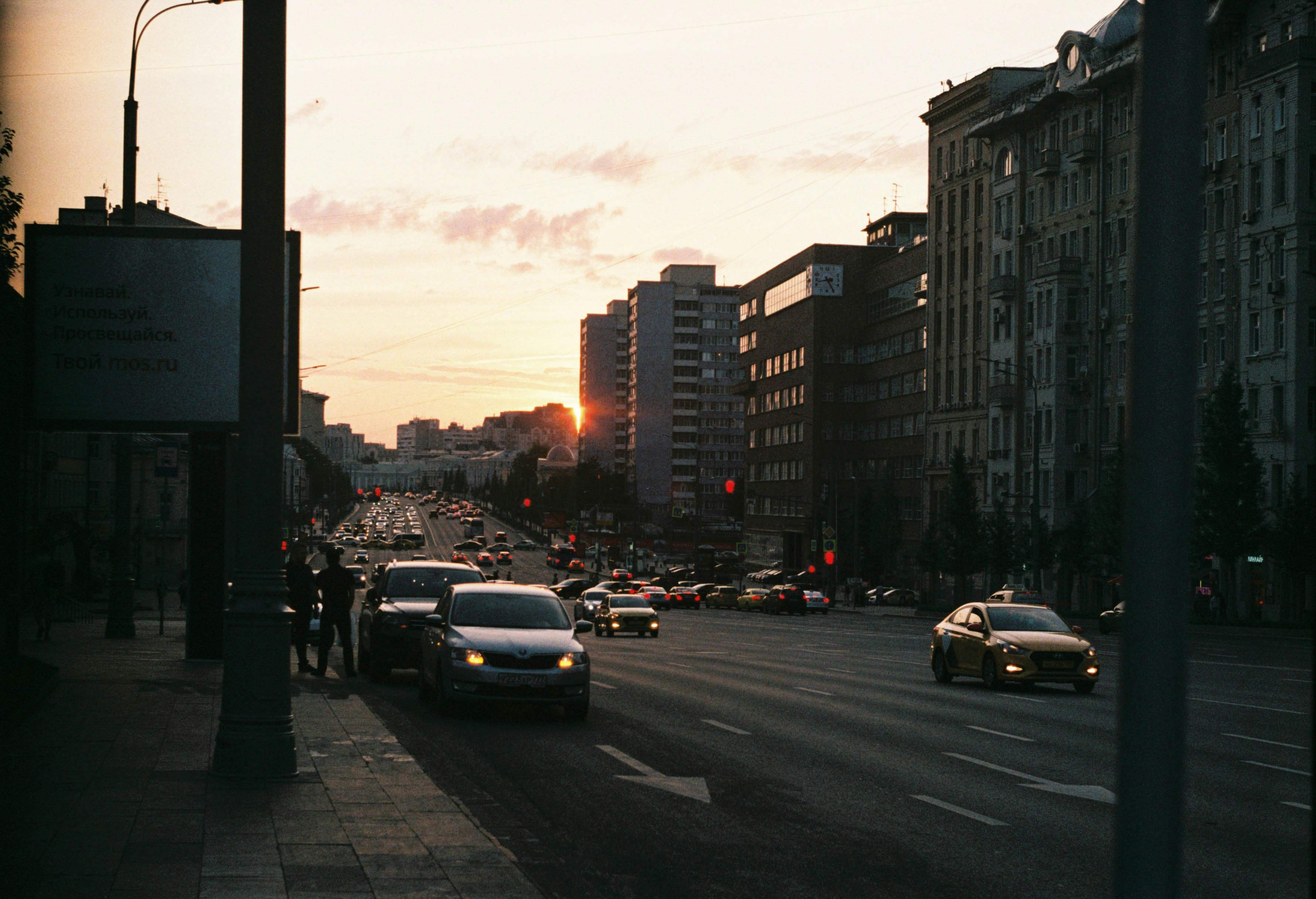 Busy city street at sunset, with cars in motion and buildings lining the road. Traffic lights and pedestrians add to the urban atmosphere.