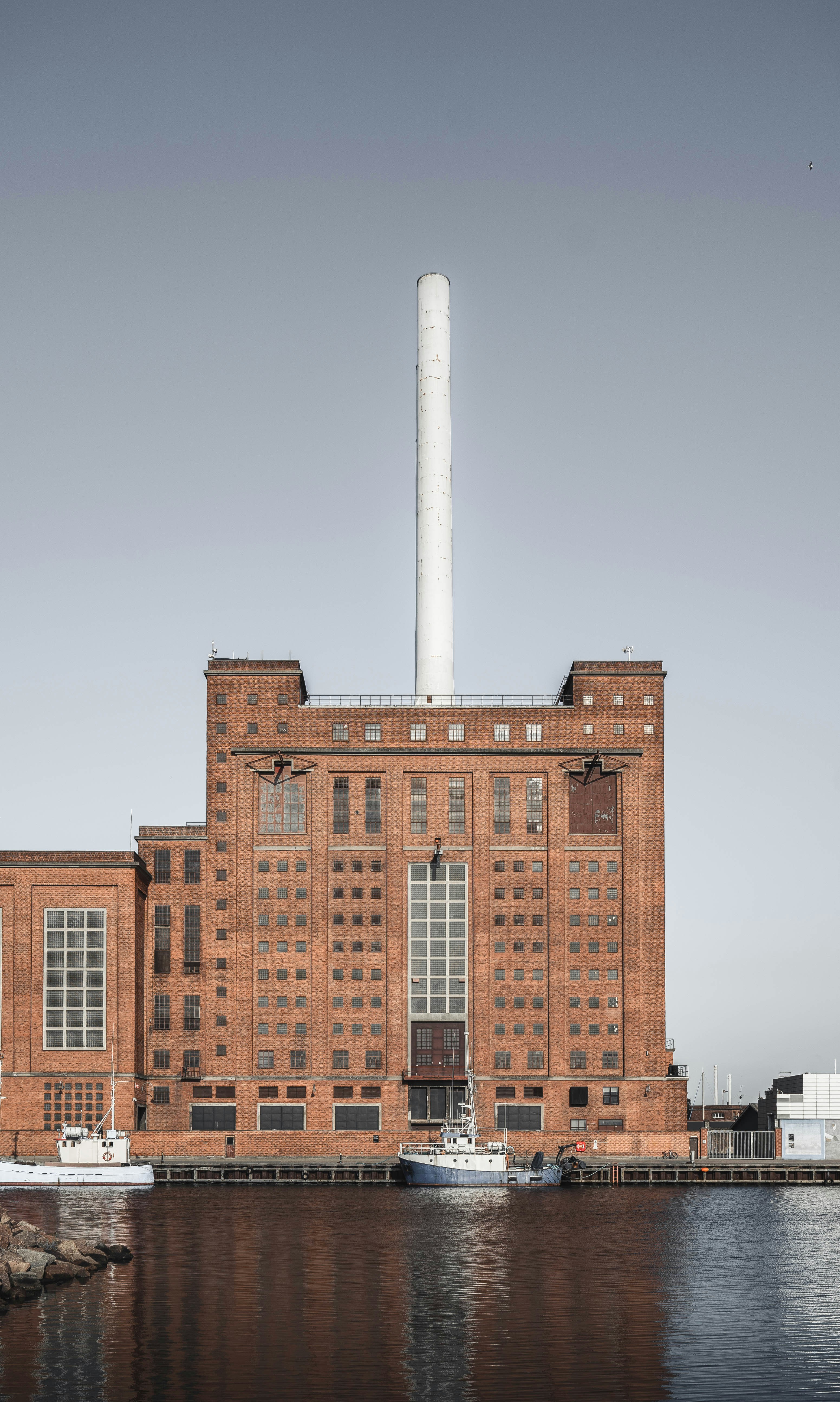 Historic brick factory building with a prominent smokestack reflected in calm waters, showcasing industrial architecture.