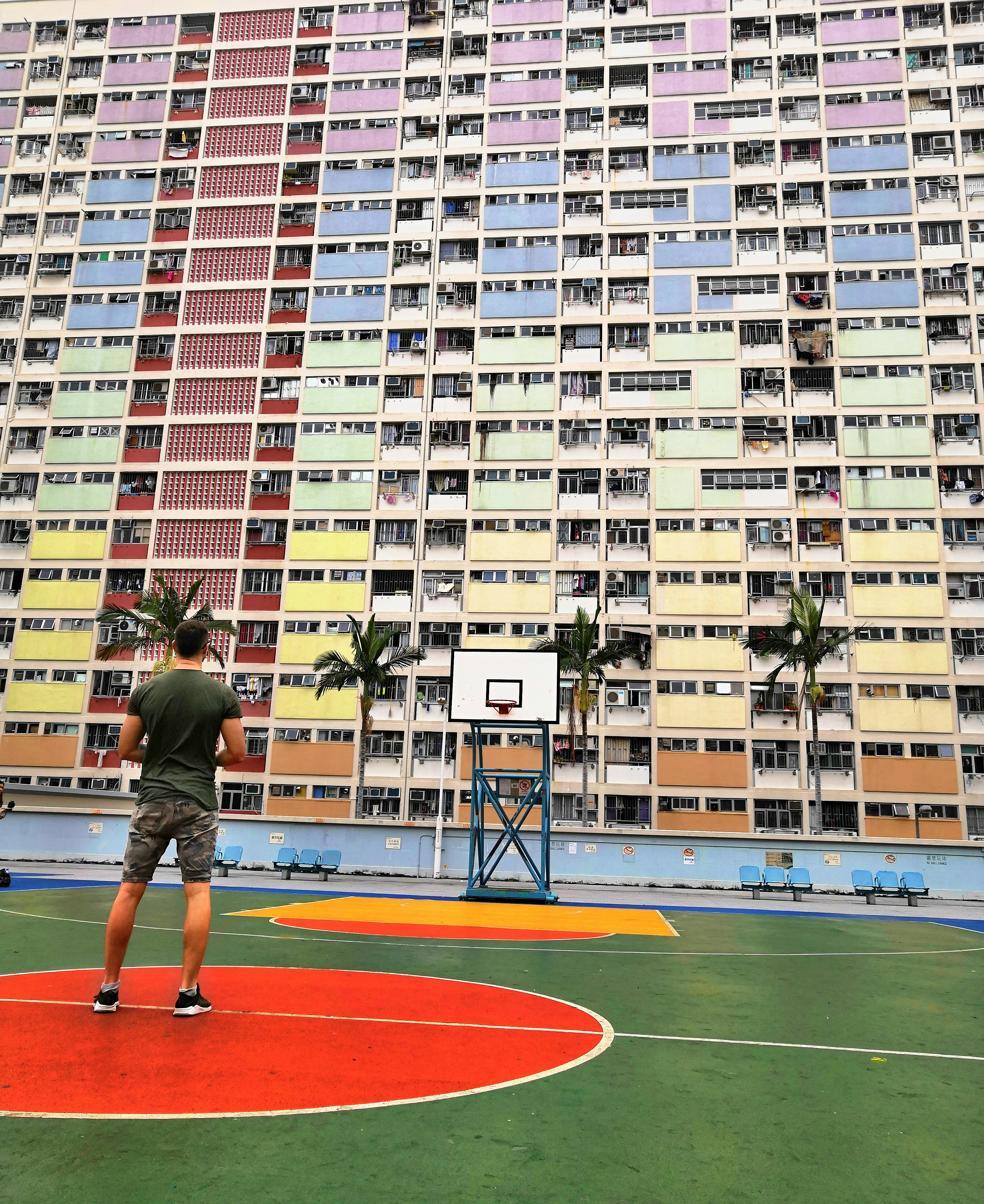 Man stands on a vibrant outdoor basketball court with a pastel rainbow apartment block towering behind; palm trees frame the scene.