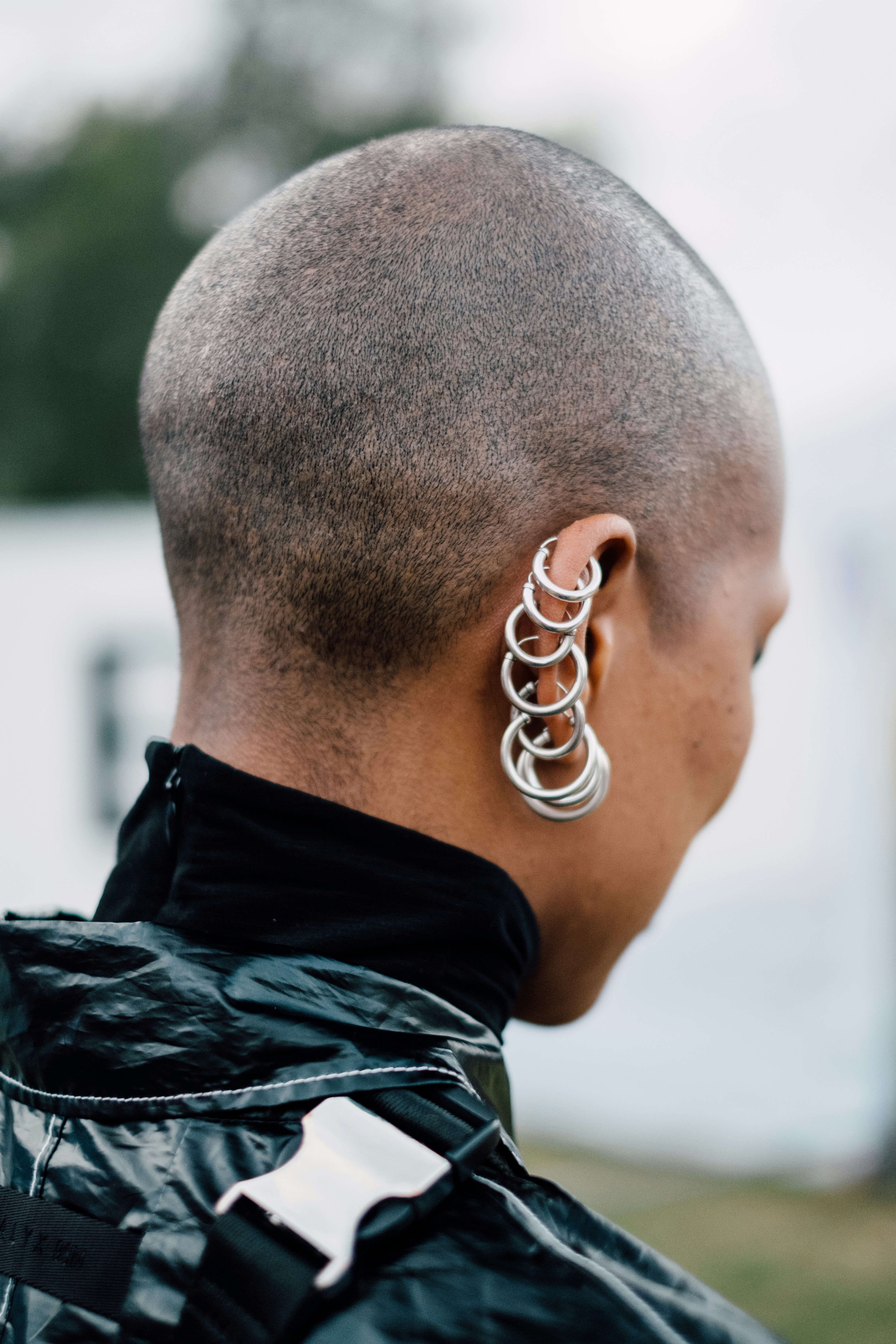 Close-up of a person with a shaved head showcasing a unique multi-ring earring against a blurred background. The focus is on the intricate design and modern style of the accessory.