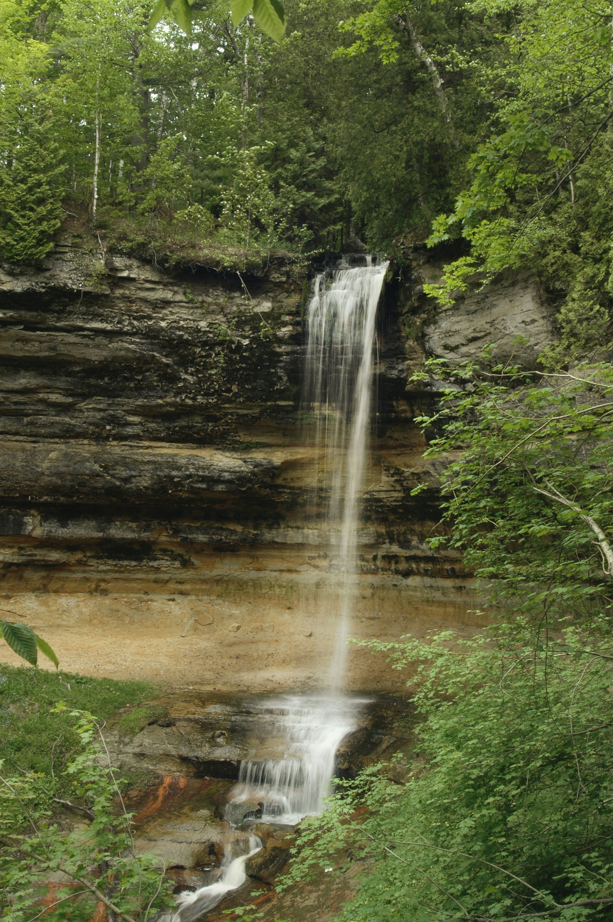 A serene waterfall descends over layered rock formations, surrounded by lush greenery. The scene captures the tranquility of nature's flow.