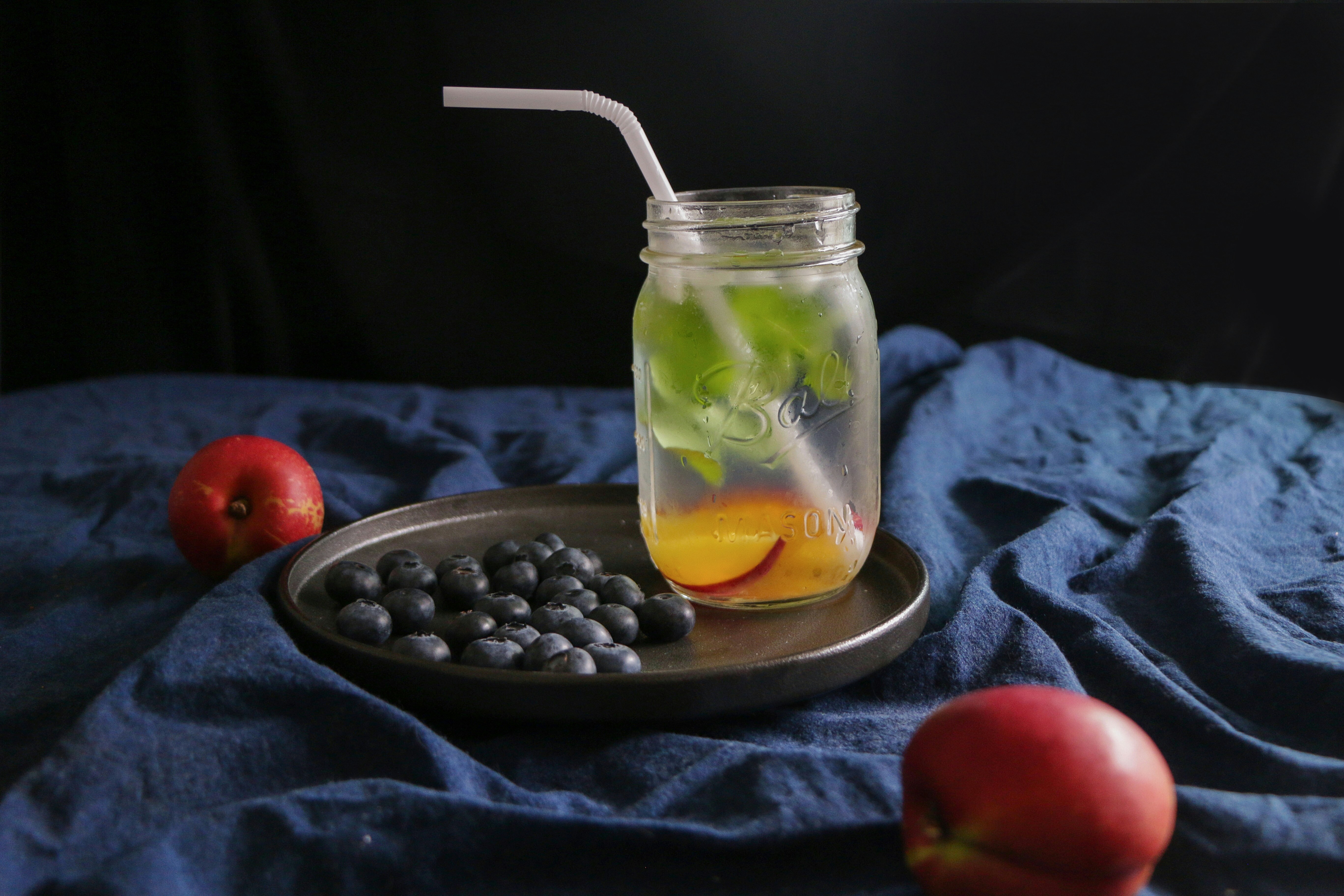 A mason jar filled with a refreshing drink featuring citrus and mint, accompanied by a plate of blueberries and red apples on a deep blue fabric backdrop.