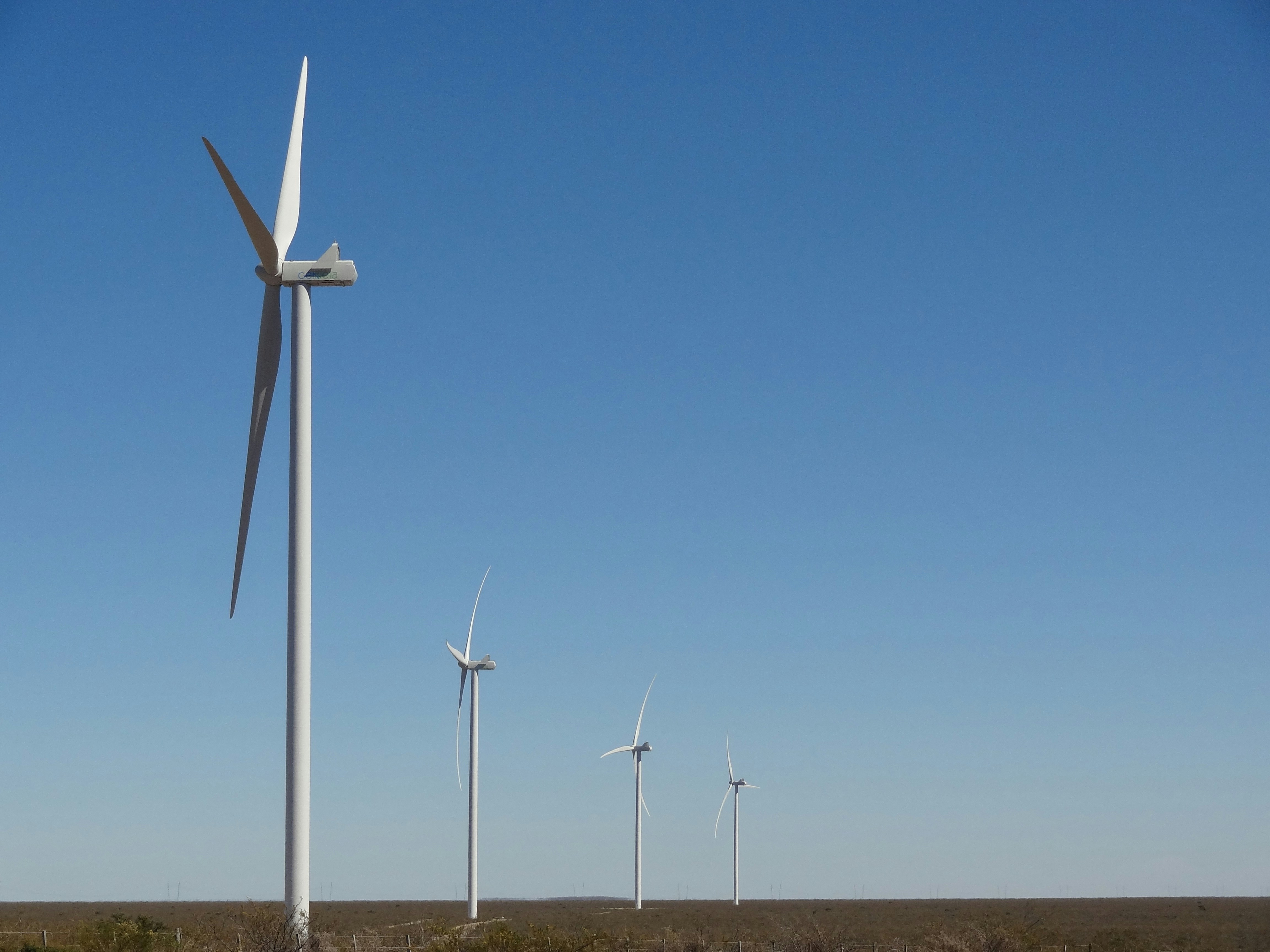 Row of wind turbines stretching toward a flat horizon under a blue sky, with a clear separation between land and sky. The scene emphasizes clean energy and vast open space.