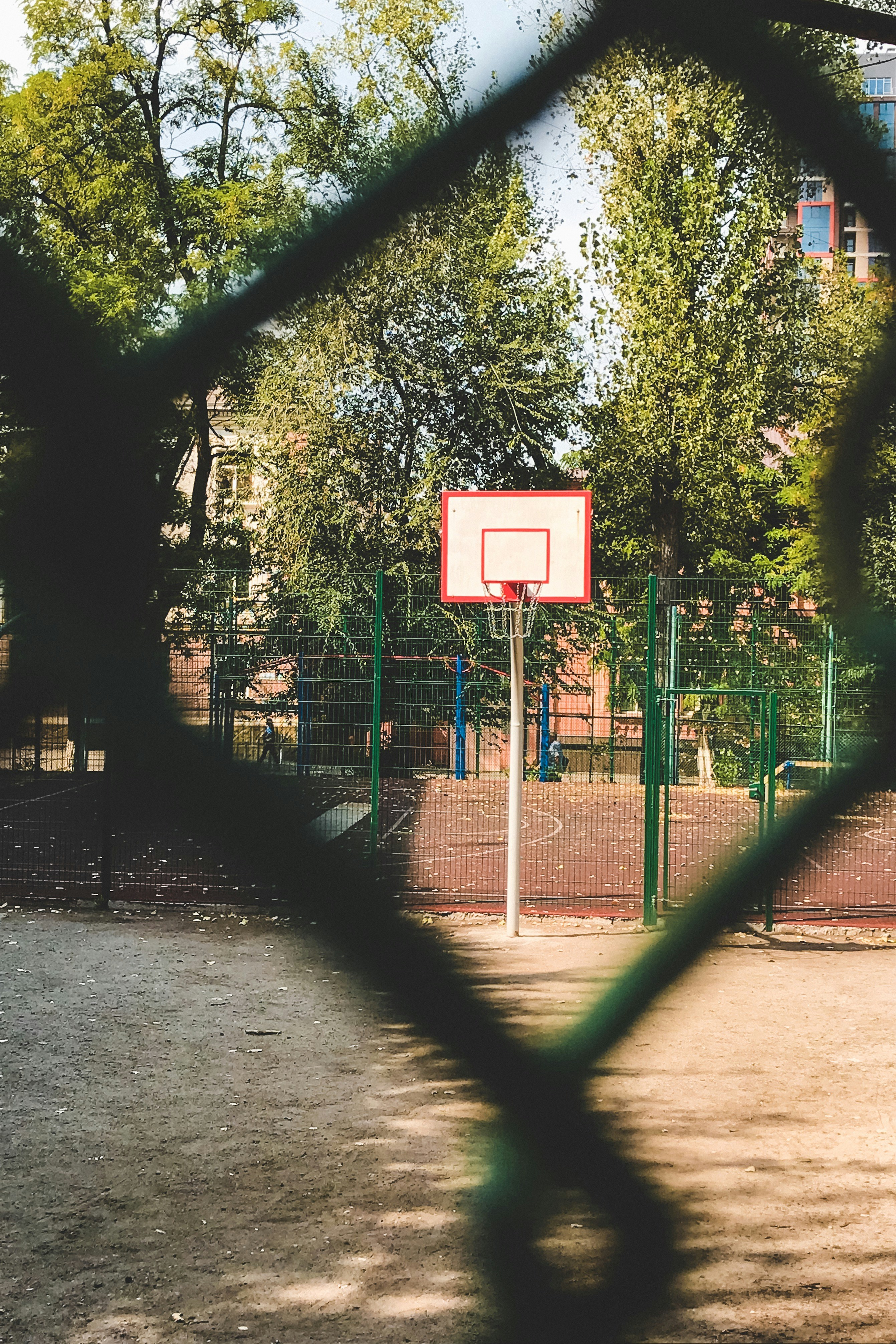 Basketball hoop framed by a chain-link fence in a playground setting, surrounded by lush trees. 