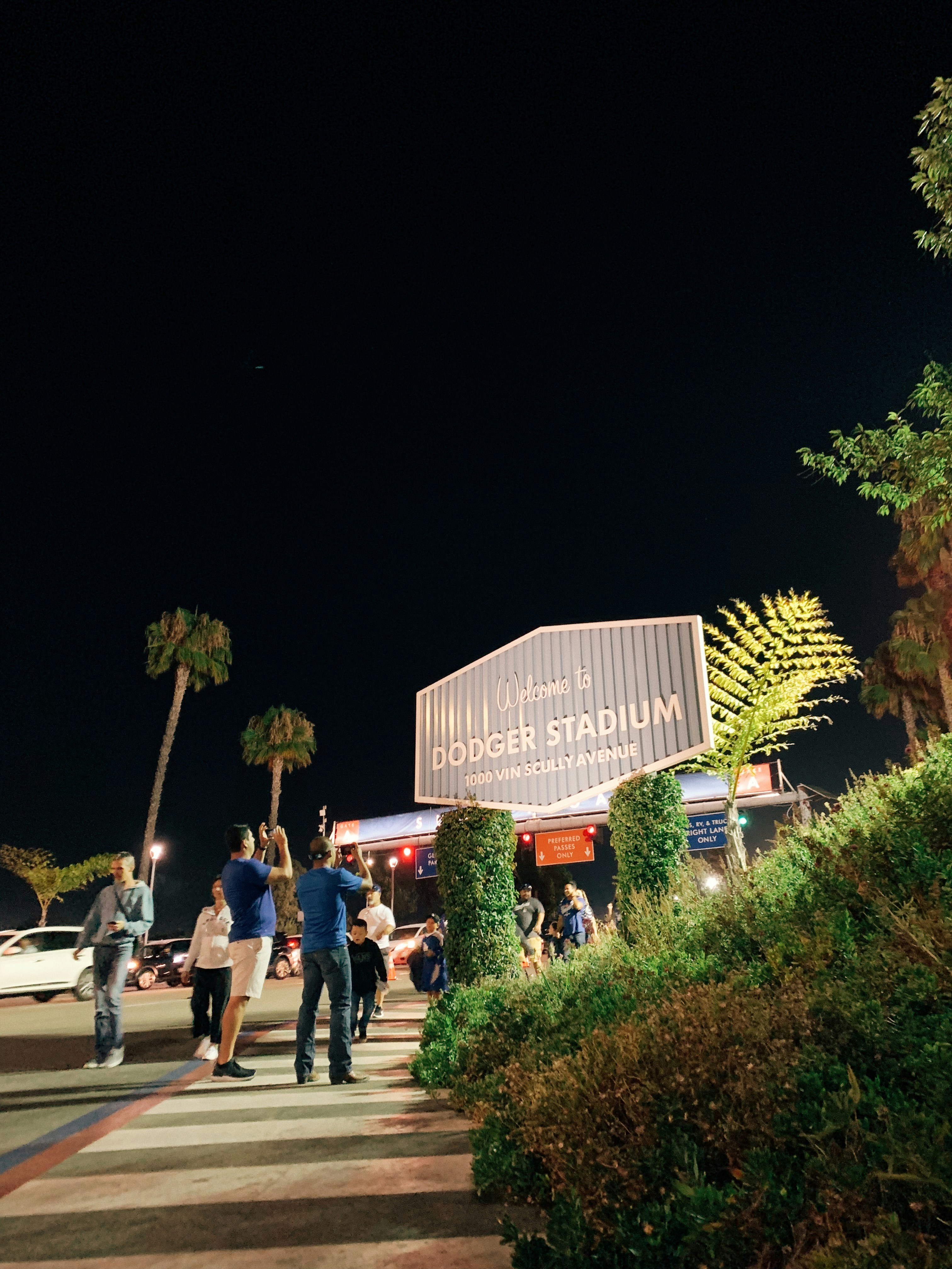 Crowd gathering under the illuminated Dodger Stadium sign at night, surrounded by lush greenery and palm trees.