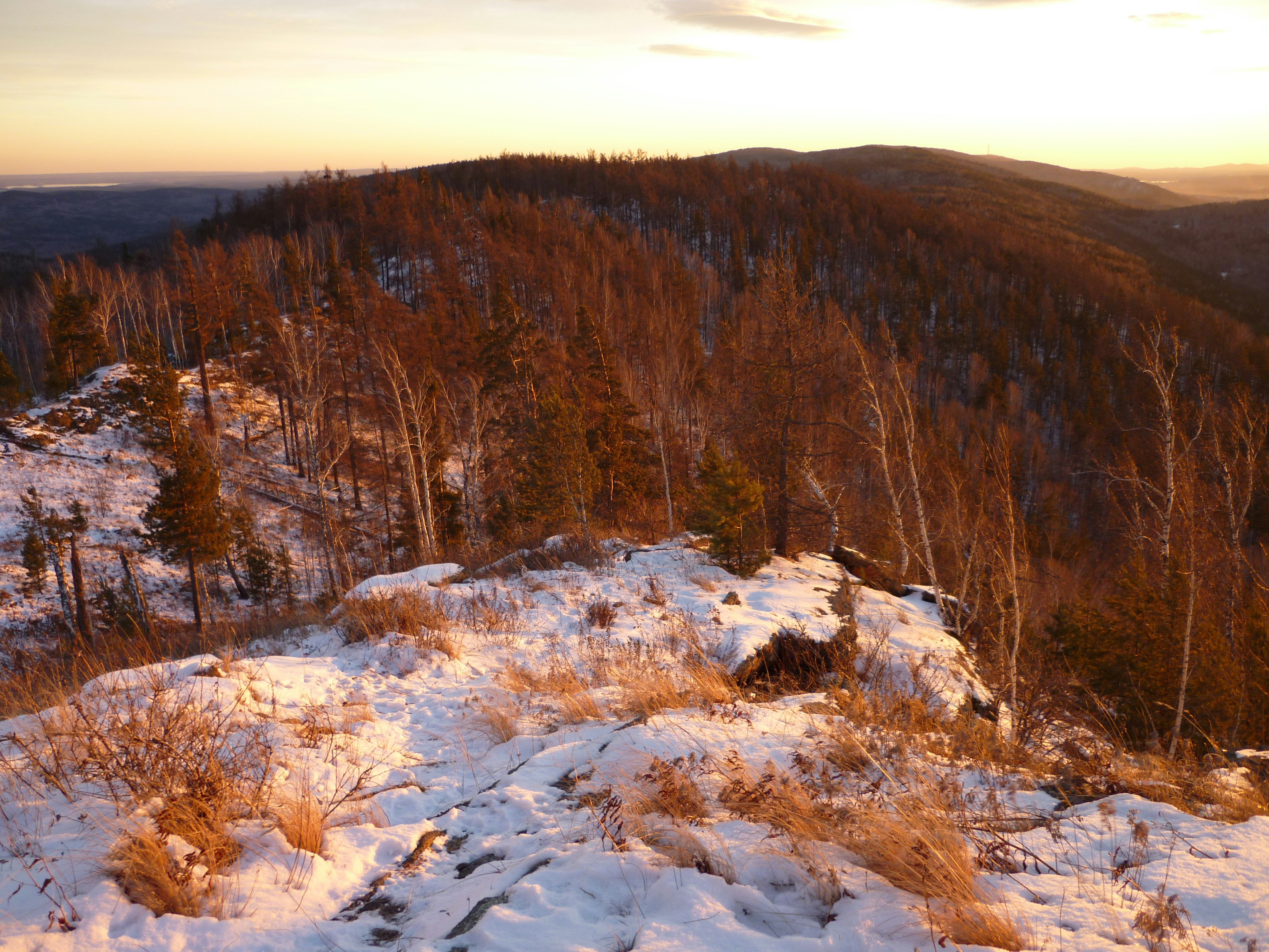 Snow-covered rocky outcrop overlooking a serene landscape of bare trees at sunset.