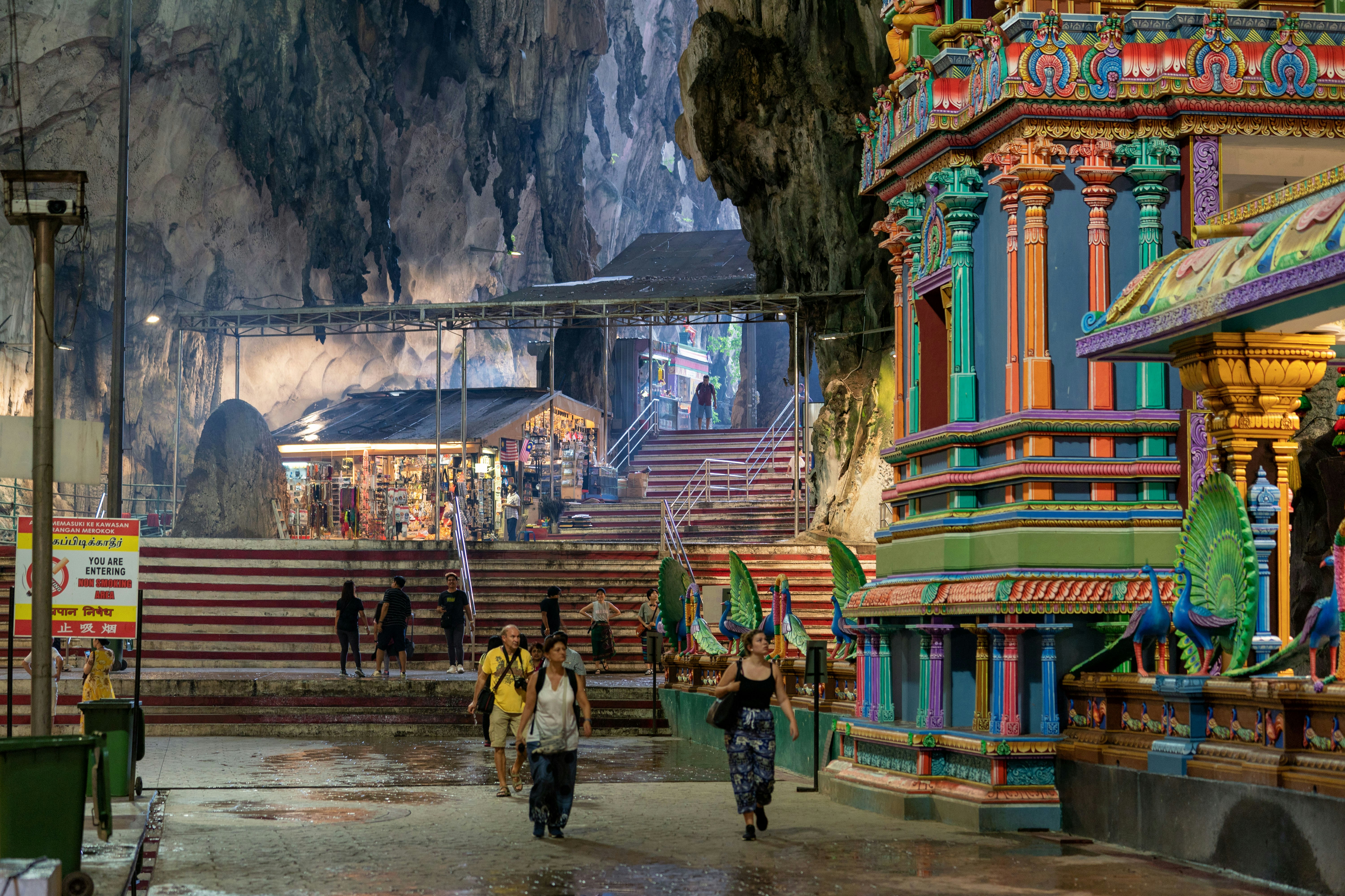 Colorful temple structures nestled within a limestone cave, with visitors exploring the vibrant setting. The scene showcases the blend of nature and culture.