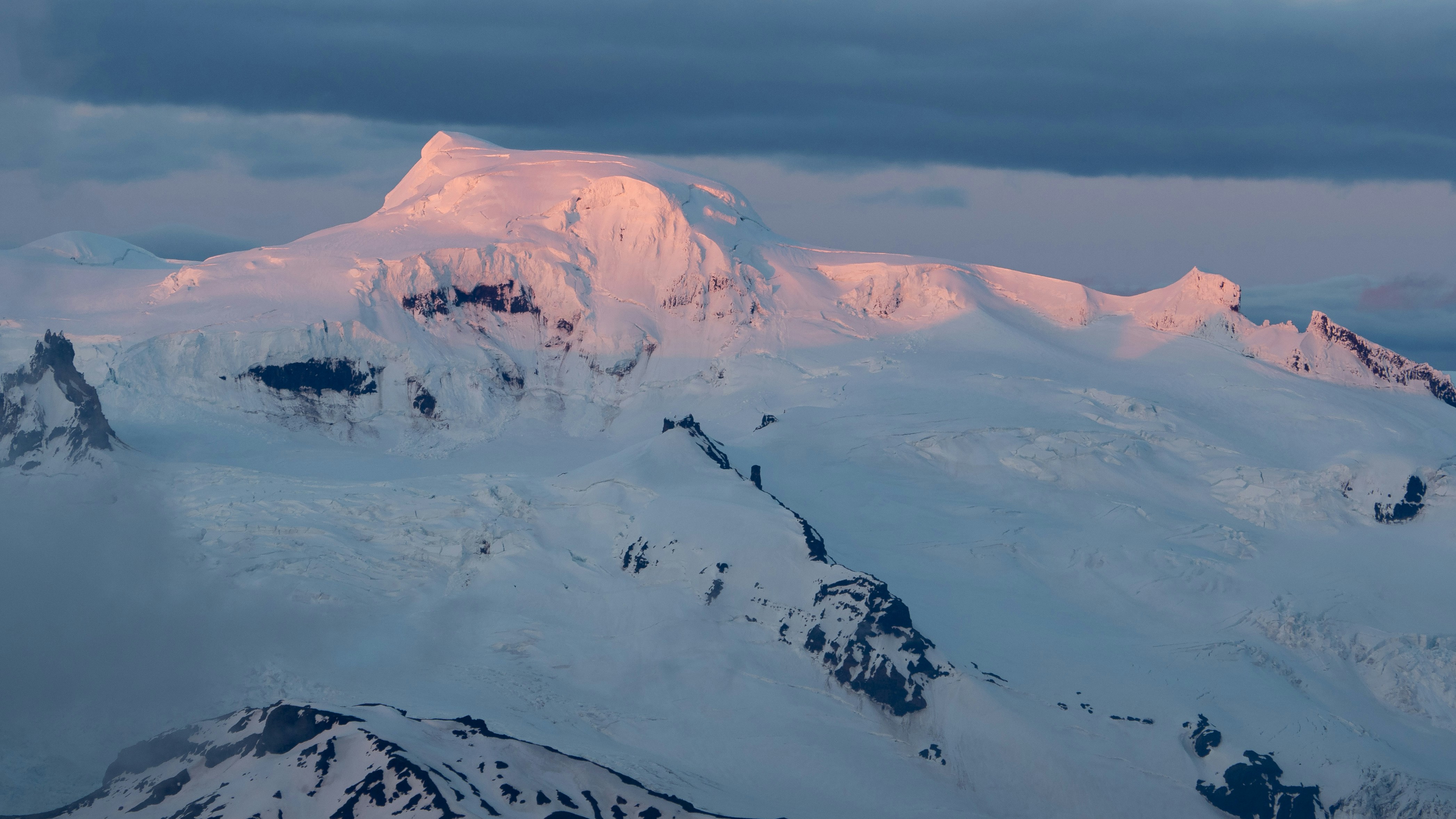 Snow-covered Alpine ridge bathed in pink dawn light, with a distant glacier peak.