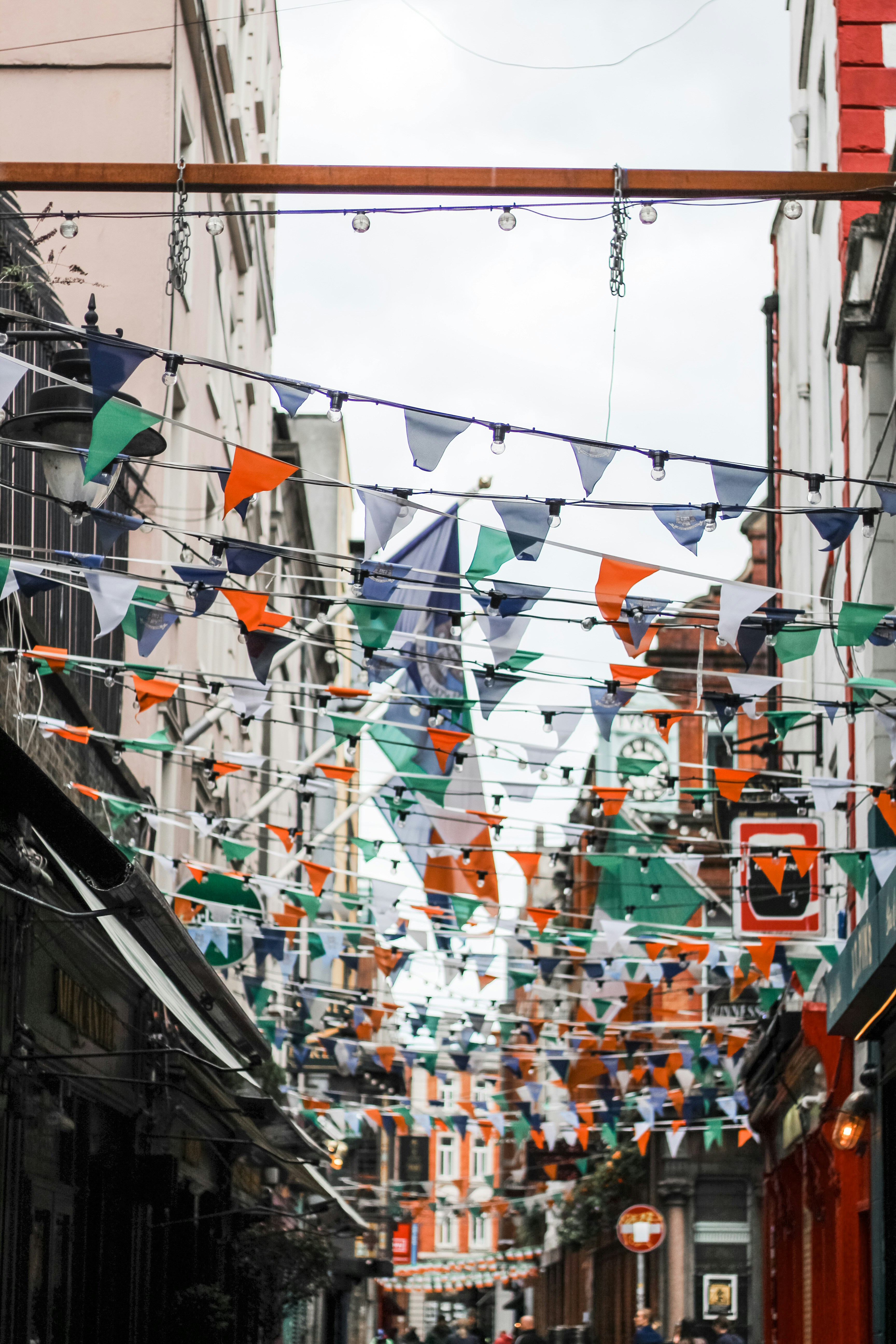 Colorful triangular flags in green, white, and orange hang across a bustling alley, adding a festive atmosphere to the urban scene.