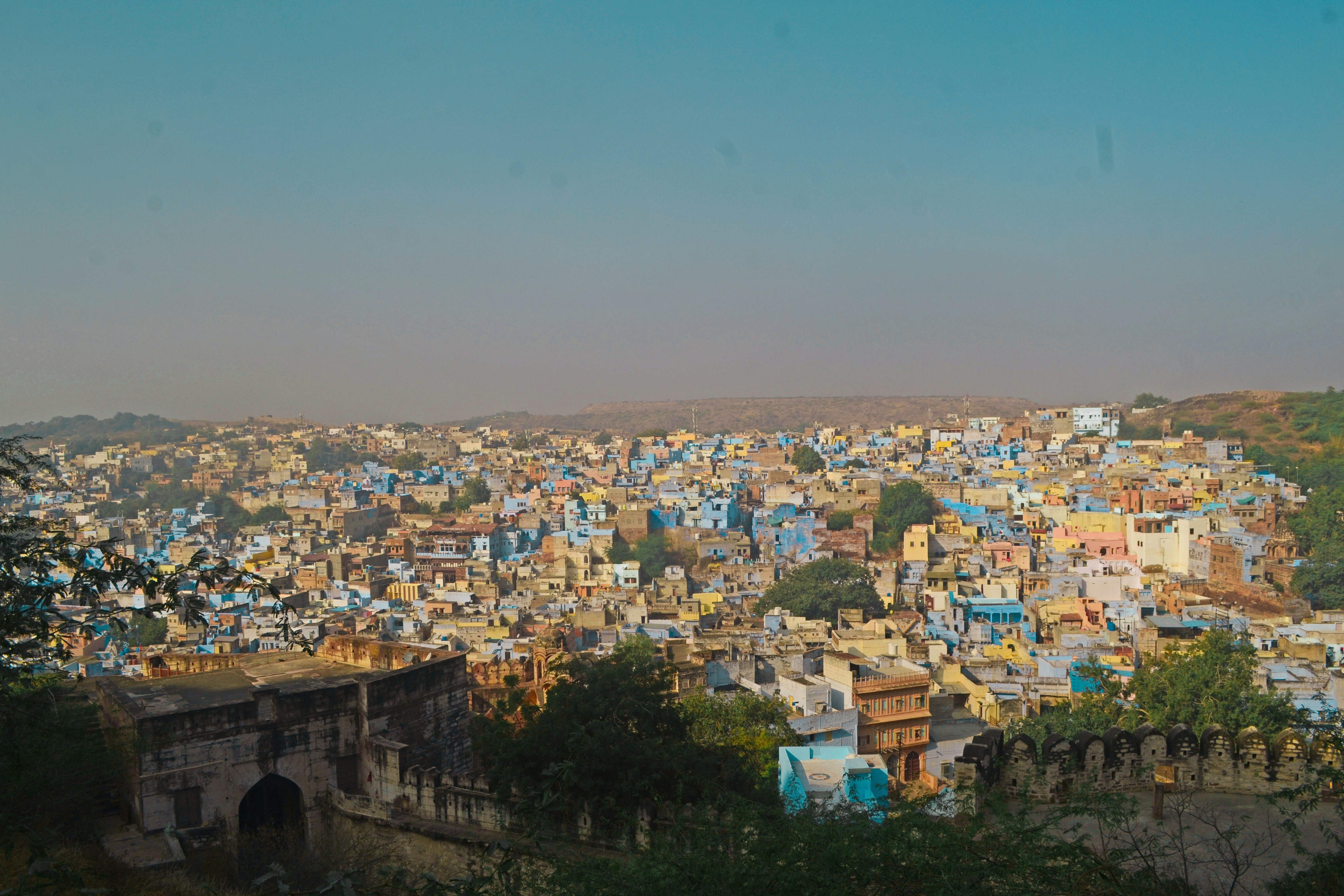 Vibrant blue houses clustered together in the city of Jodhpur, showcasing the unique architectural style of the region. Lush greenery and distant hills frame the scene.