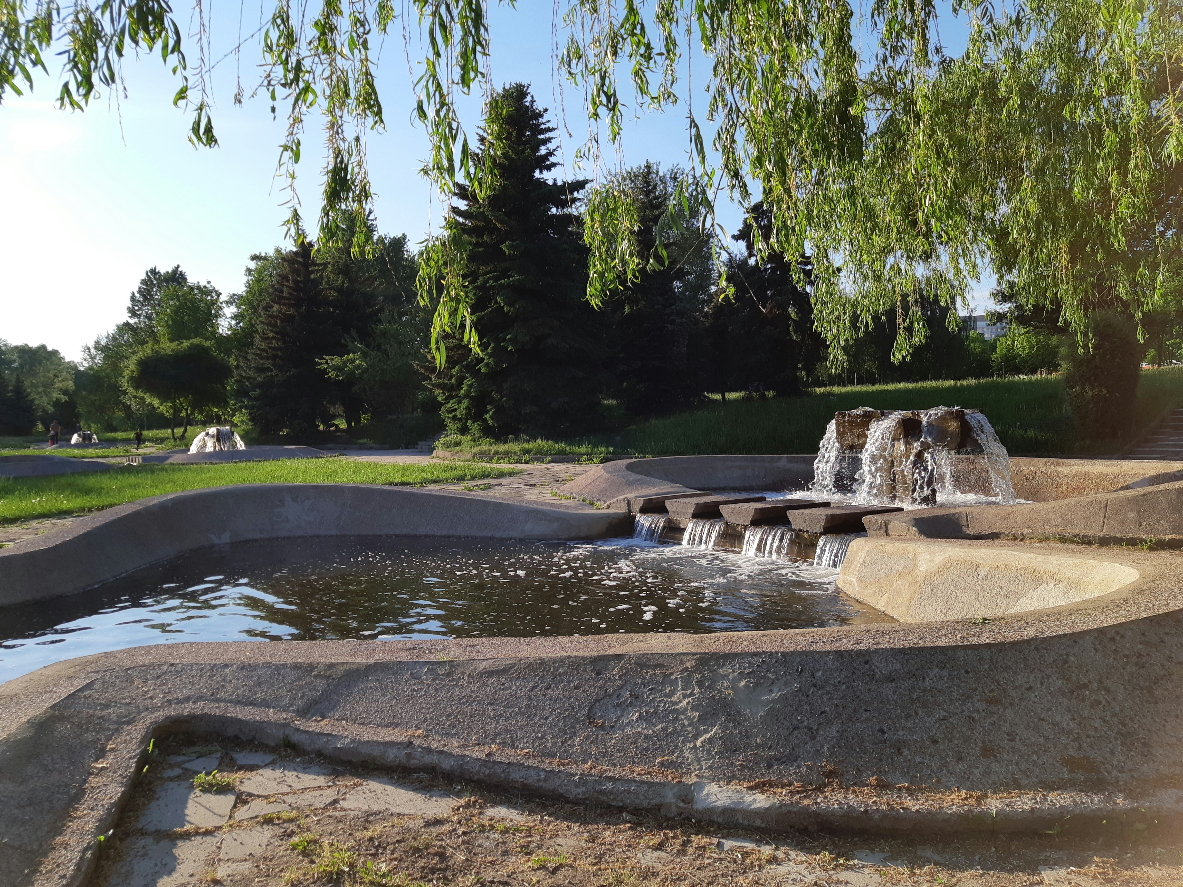 Small waterfall cascading into a serene pond surrounded by lush greenery on a sunny day.