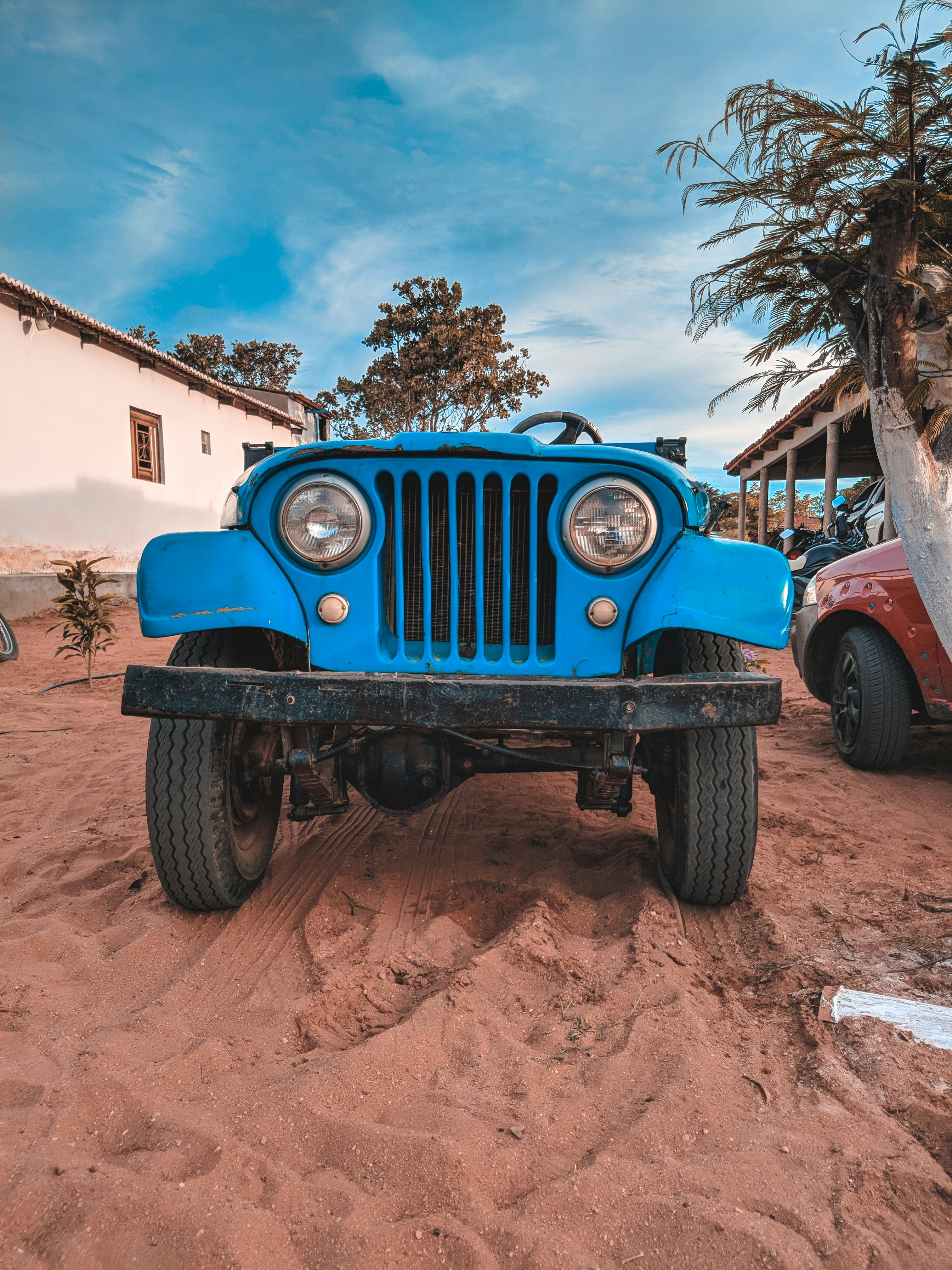 An iconic blue vintage jeep parked on sandy ground, surrounded by lush greenery and rustic buildings.