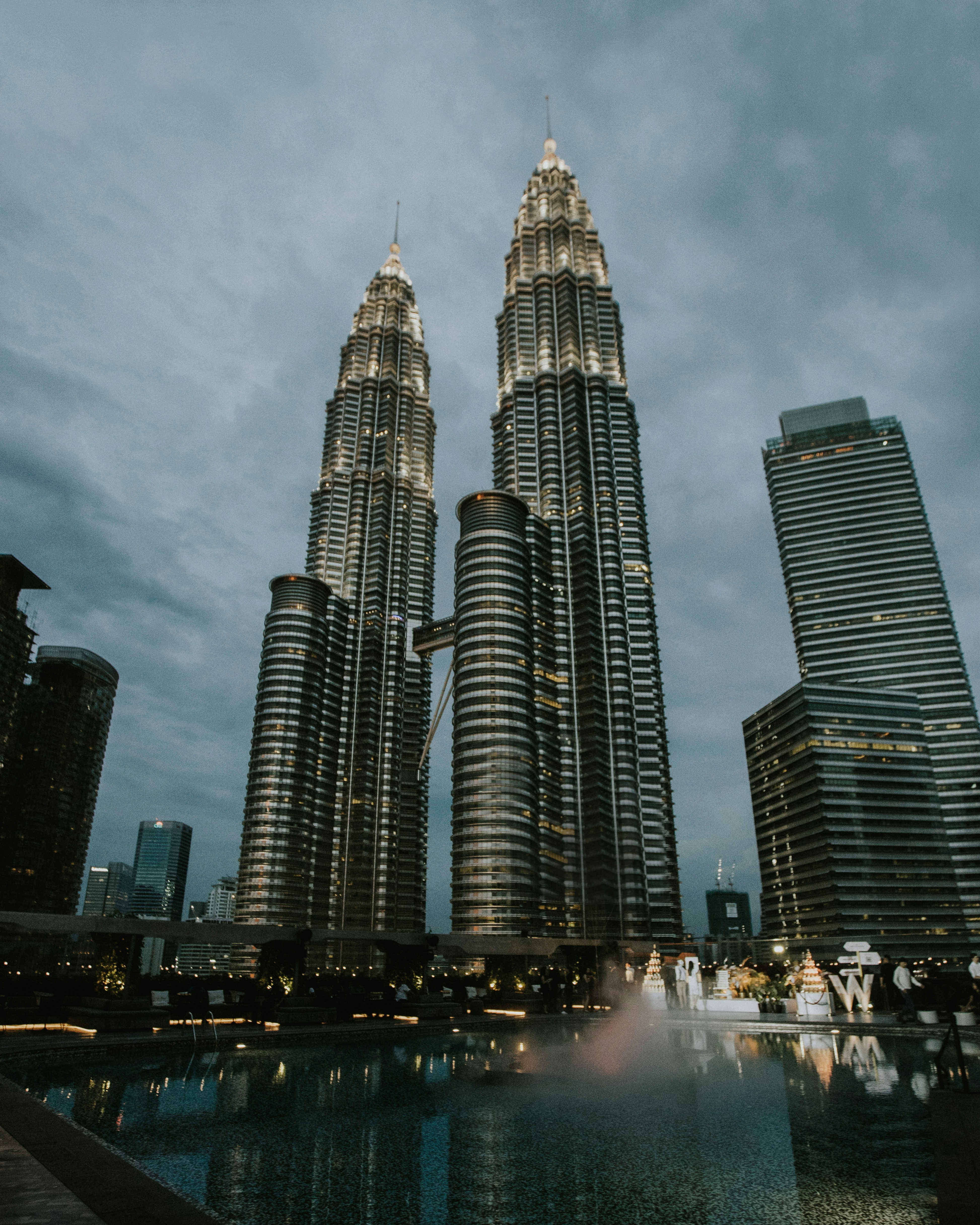 The iconic Petronas Towers rise majestically against a moody sky, reflecting in the tranquil waters below.