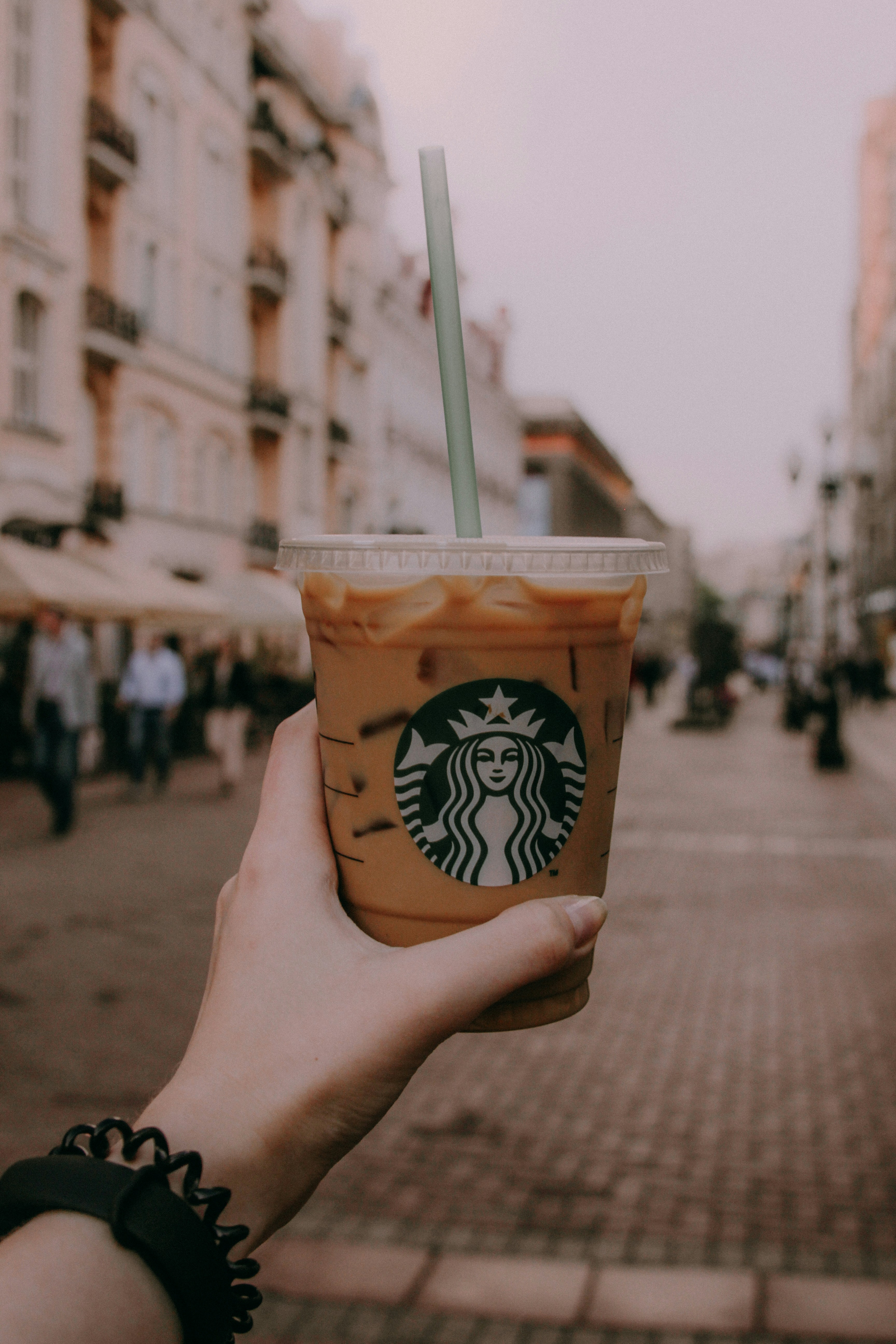 Hand holding a Starbucks iced coffee against a bustling urban backdrop, capturing a lively street scene.