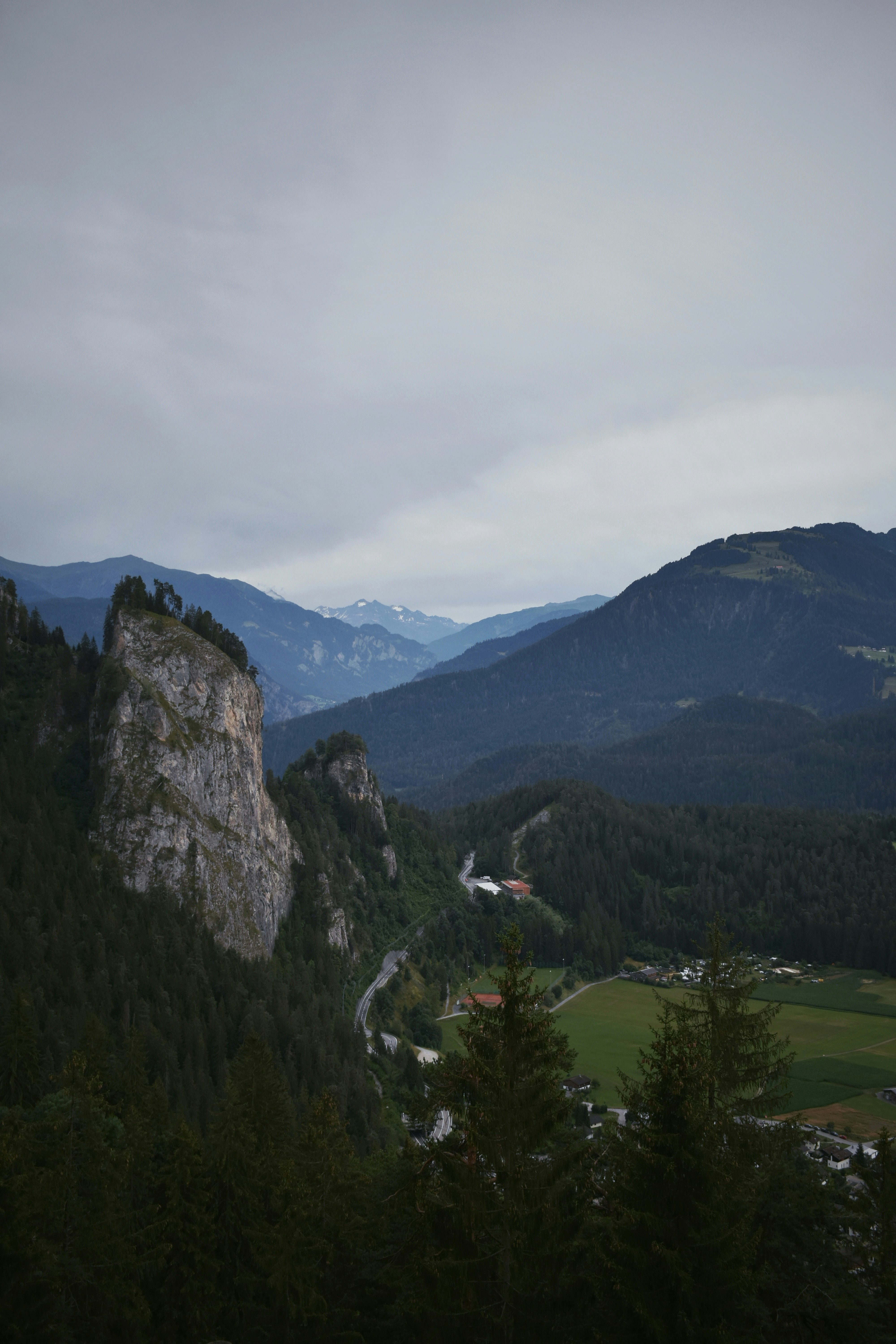 Majestic rocky outcrops rise above a lush valley, with winding roads and distant mountains under a moody sky.