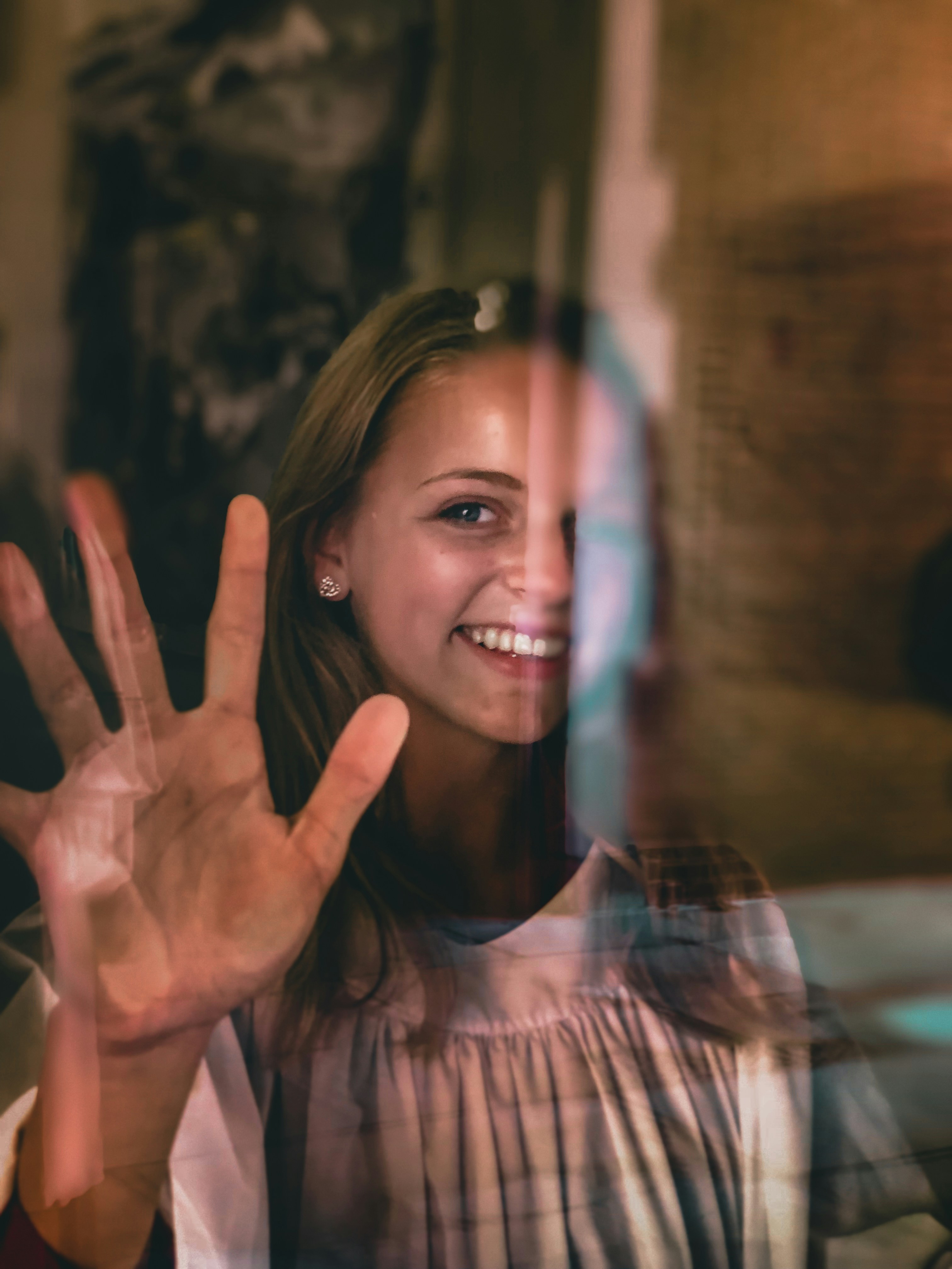 Smiling young woman with hand pressed against a glass window, creating a layered reflection effect.