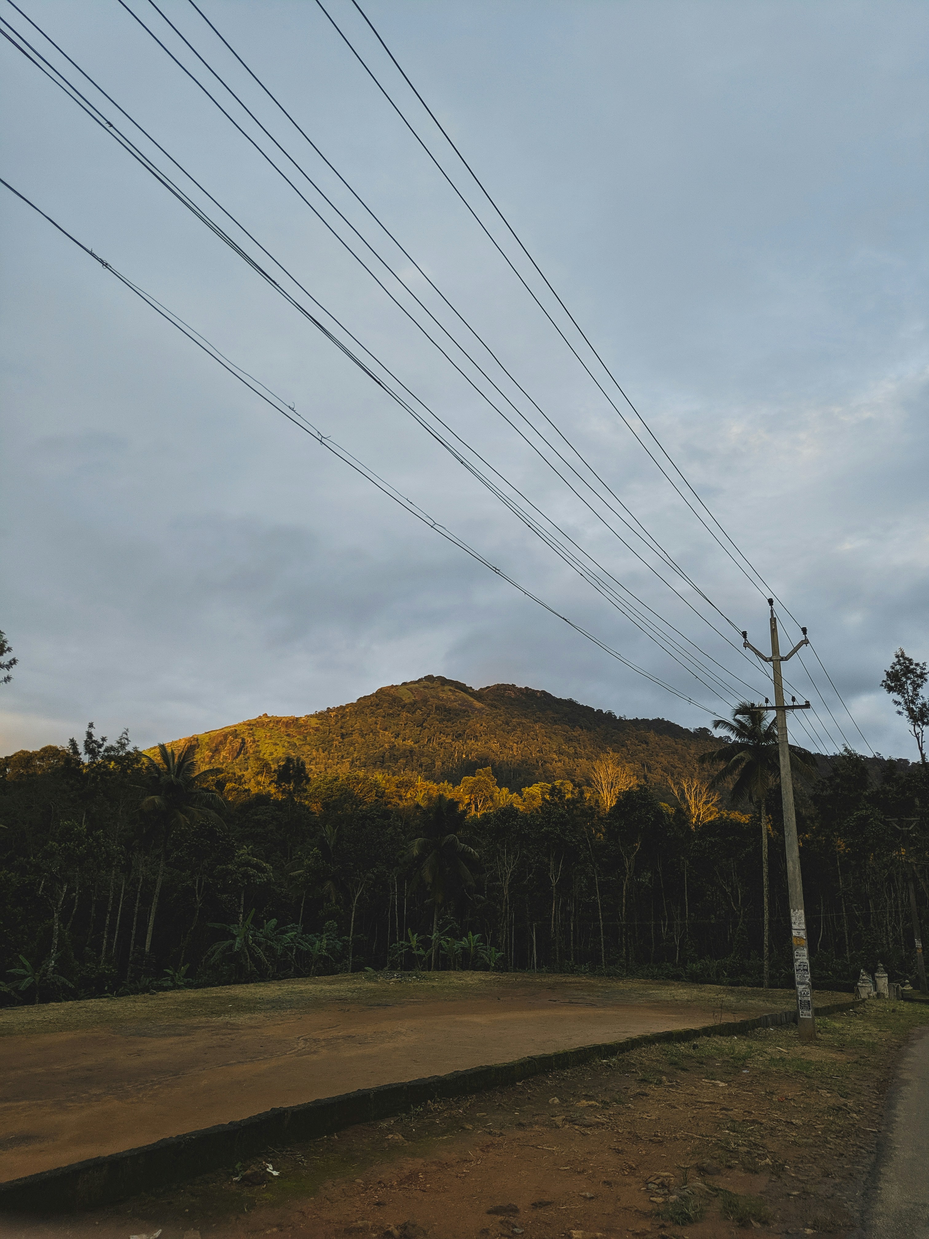 Sunlight illuminating the peak of a mountain surrounded by lush greenery and power lines against a cloudy sky.