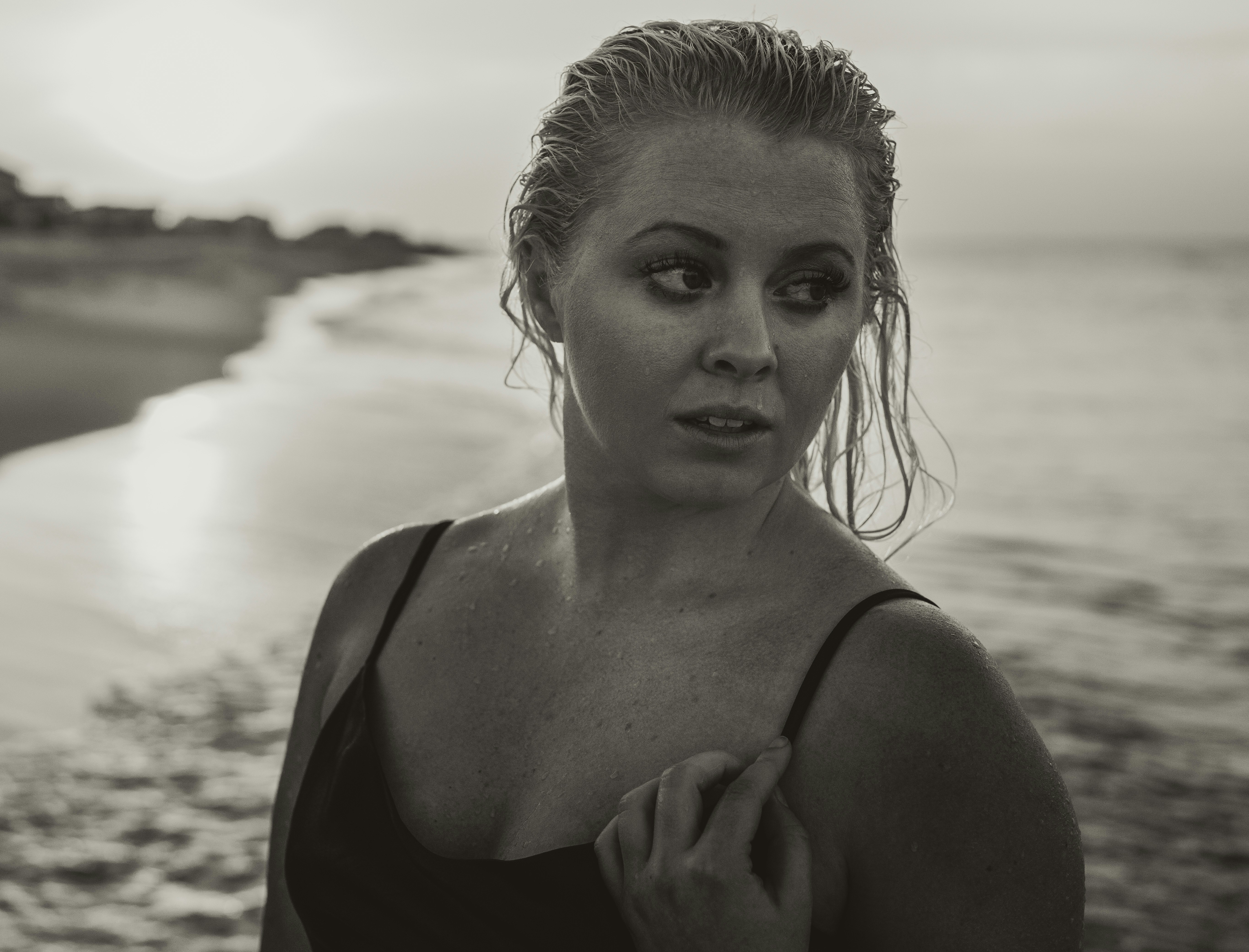 A contemplative figure stands at the water's edge, the soft waves caressing the sandy beach as the sun sets in the background.